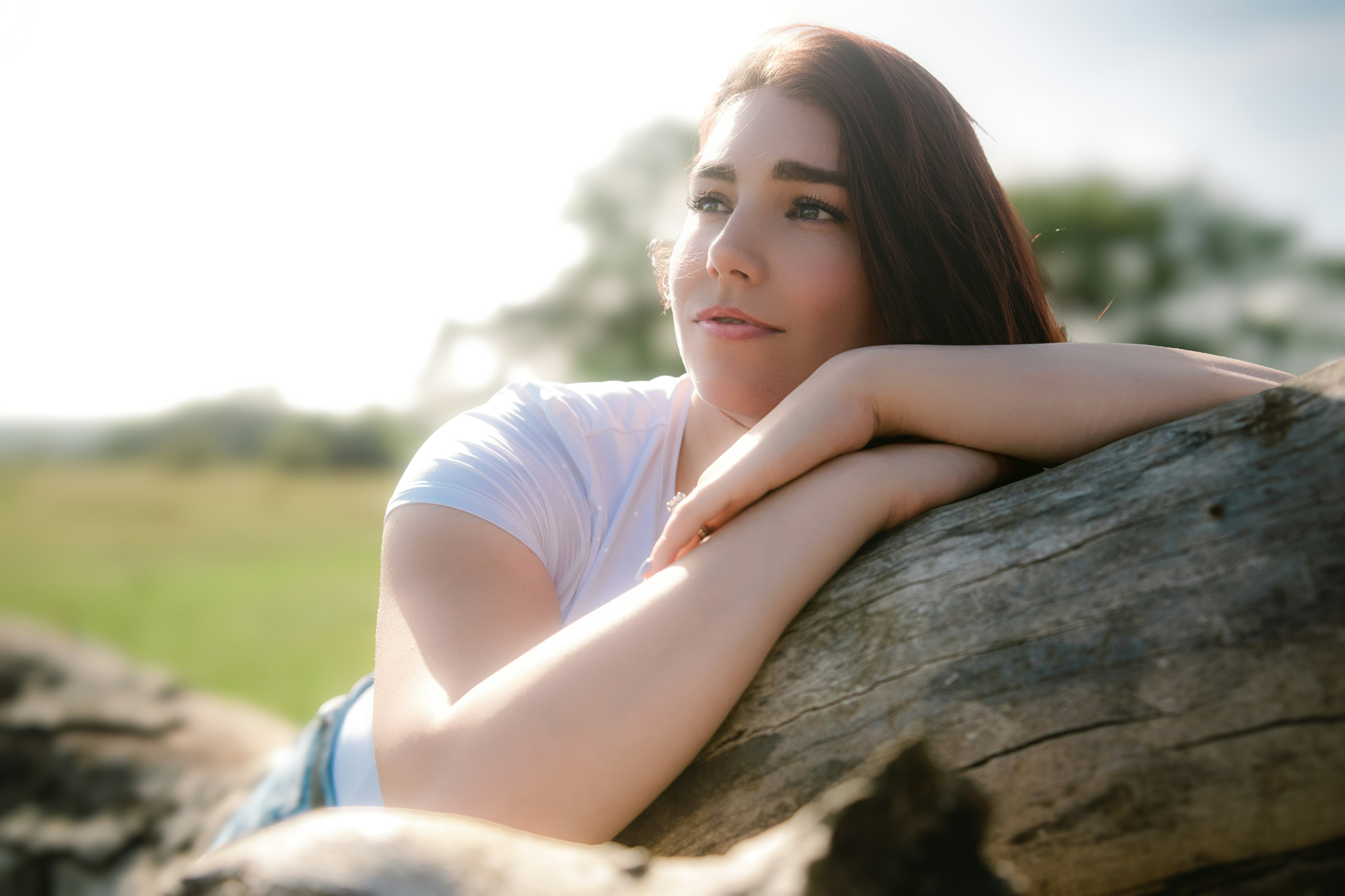 Young woman resting her arms on a weathered log, gazing into distance as bright backlight creates a warm glow; scene features natural outdoor setting with soft bokeh and a logo-free composition.