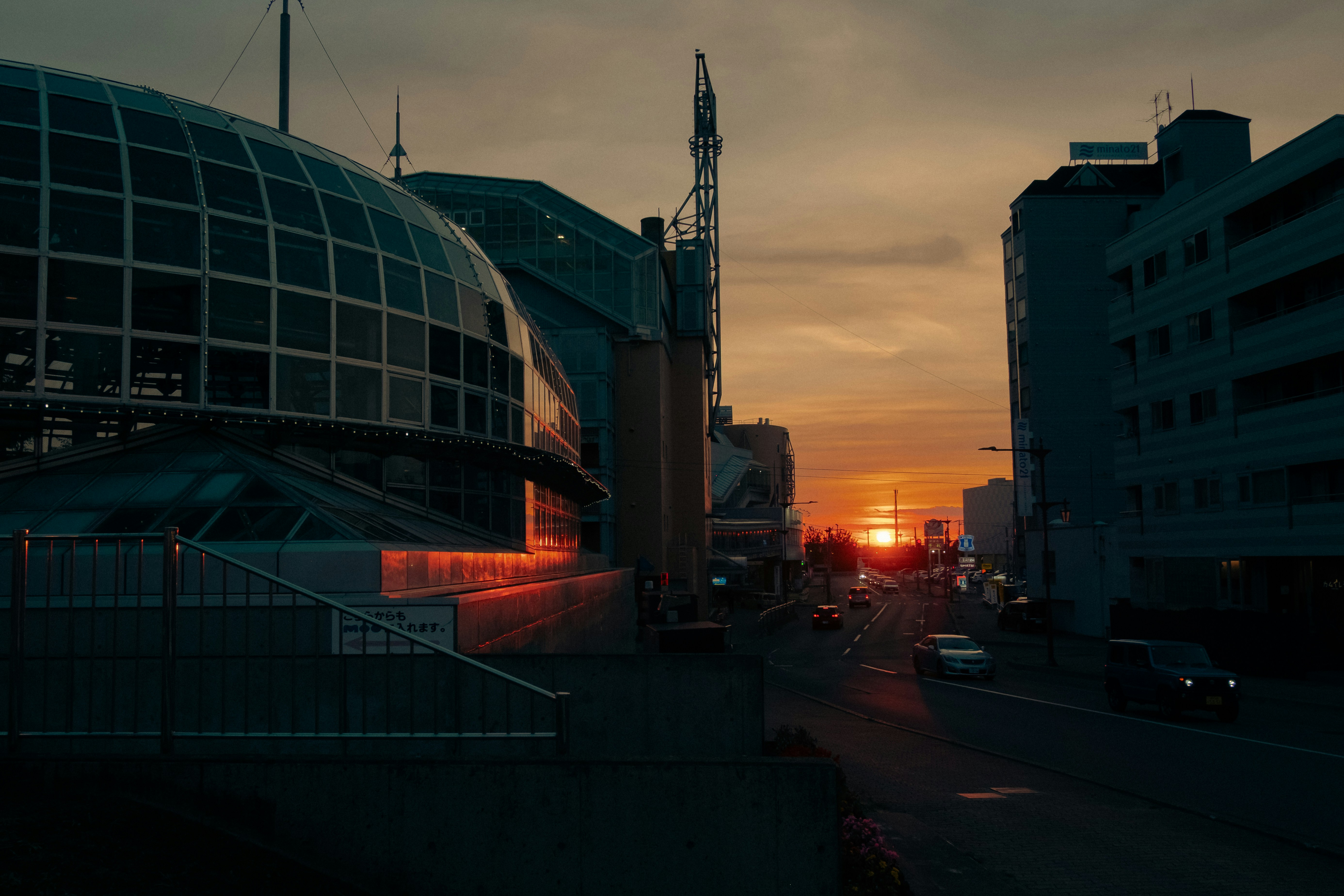 Dusk-lit city street with a glass-arched building on the left and a row of structures fading into the horizon; vehicles and warm sun low in the sky create a moody, directional scene. (Logos/architecture/urban landscape).