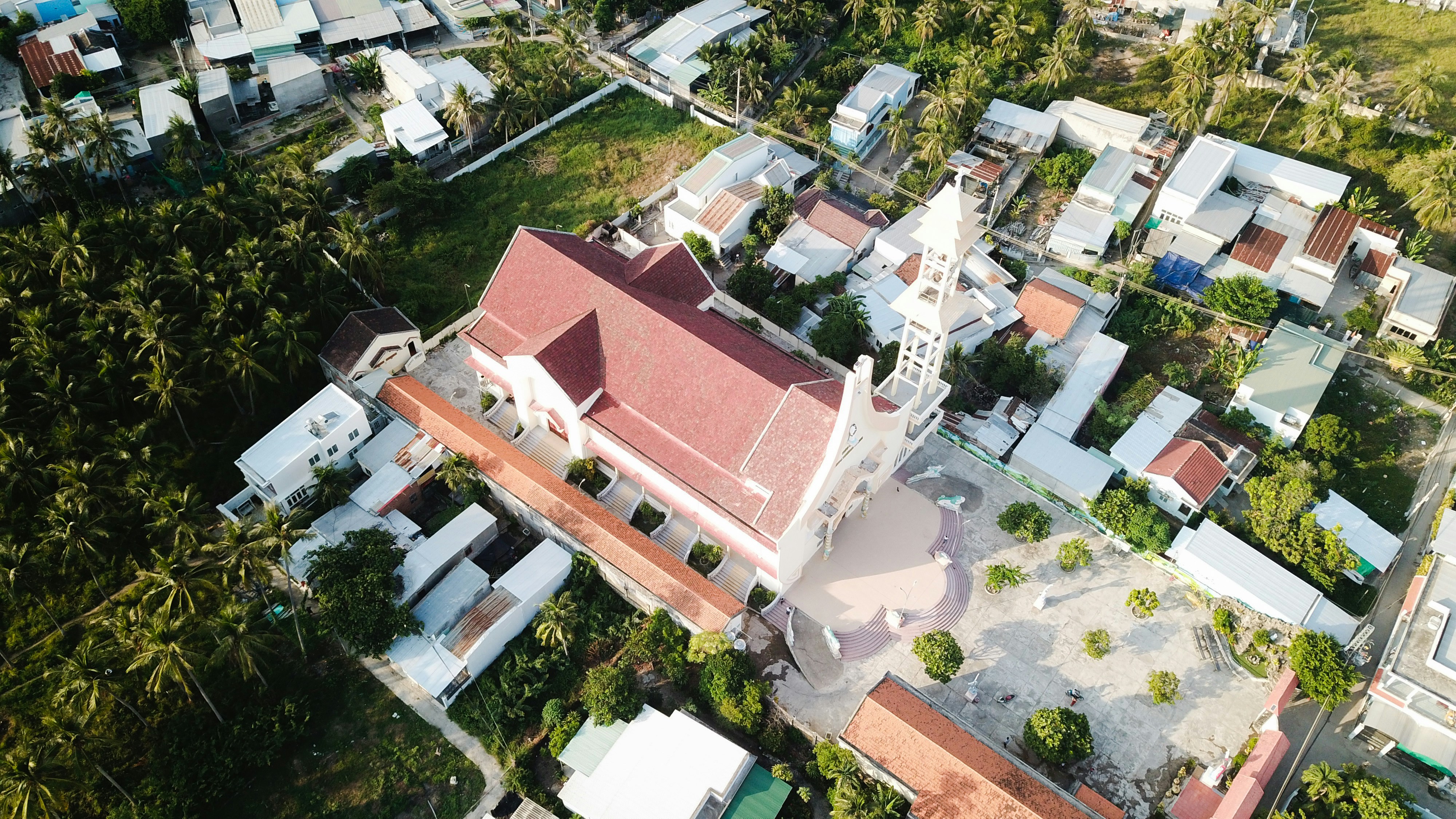 Aerial view of a red-roofed church surrounded by colorful houses and palm trees, captured from above to show the layout of the neighborhood. (This image is a drone photo.)