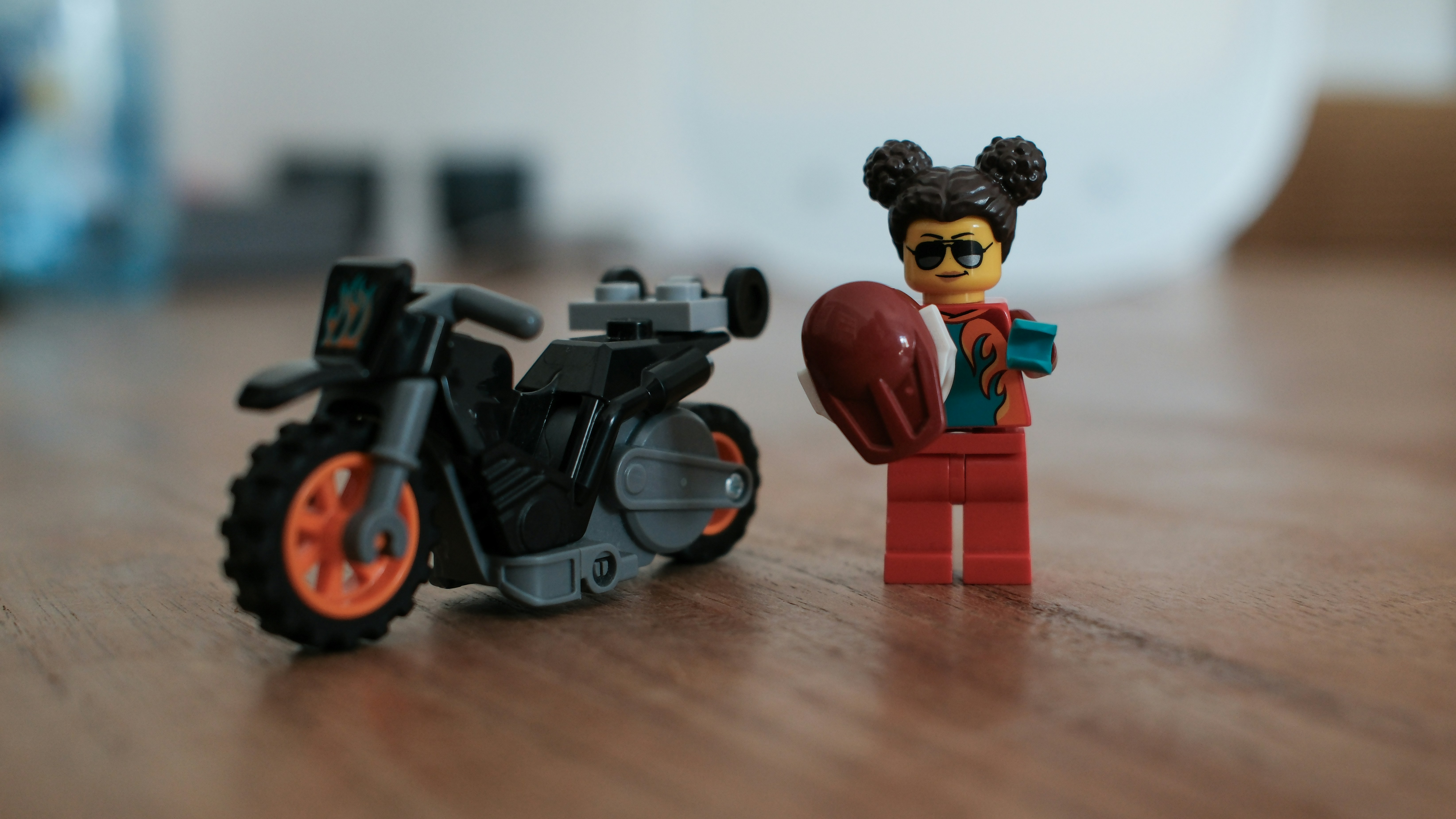 A small LEGO figure stands beside a toy motorcycle on a wooden surface, with a shallow depth of field isolating the subjects from the blurred background.