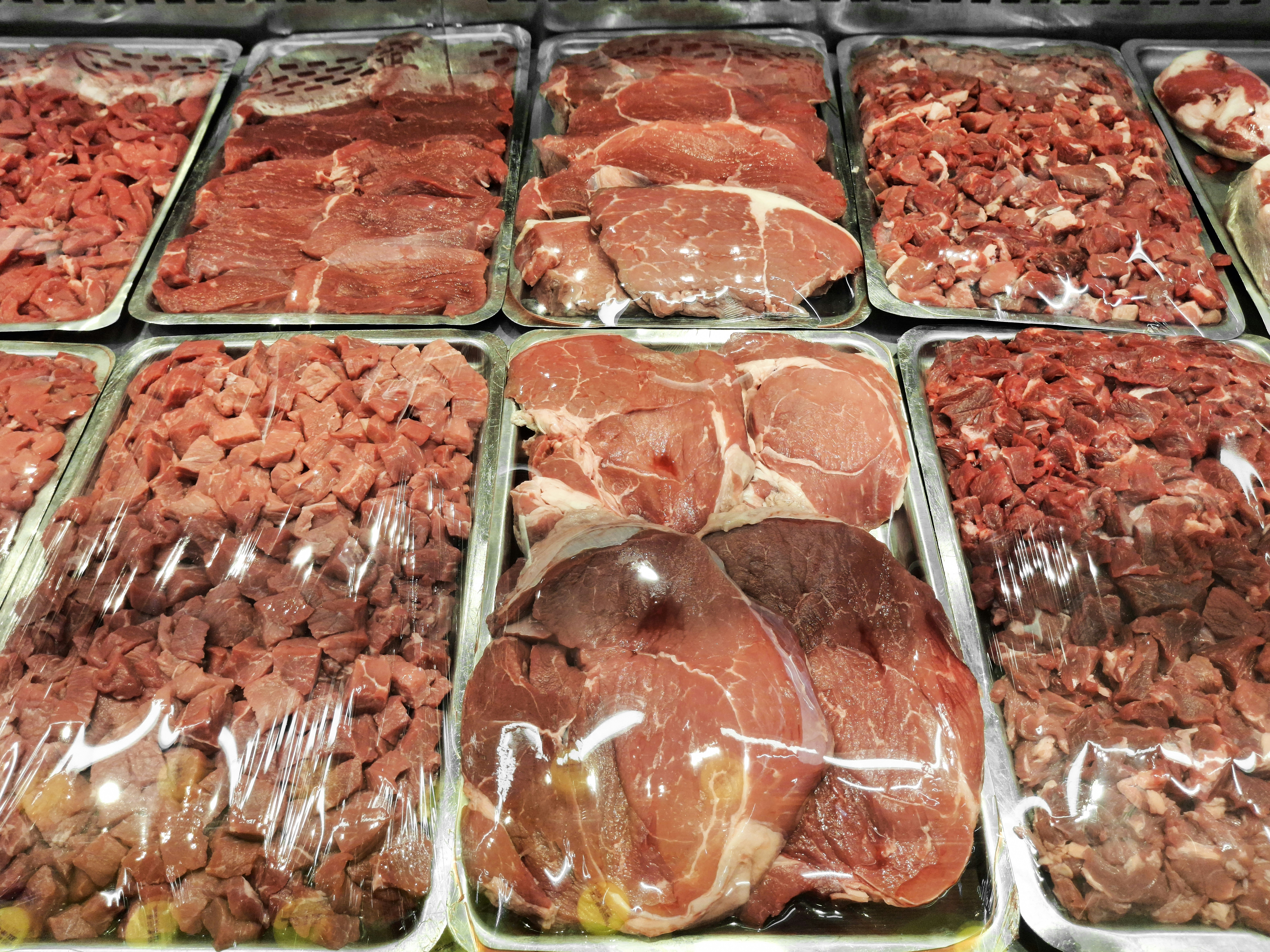 Assortment of raw cuts arranged in metal trays under plastic wrap in a market display.