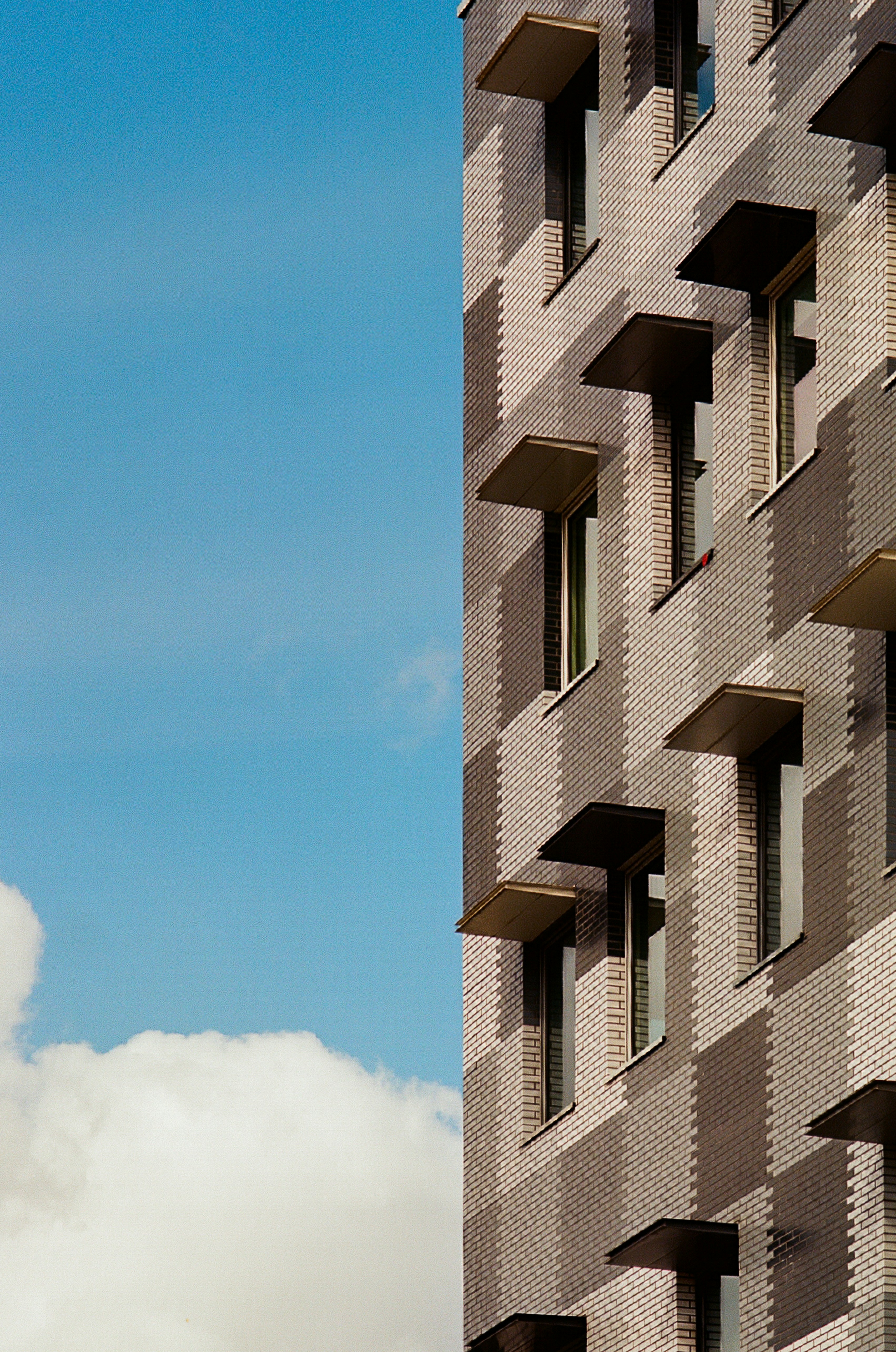 Brick building facade with projecting balconies against a blue sky with a single cloud, negative space composition, color film photography.