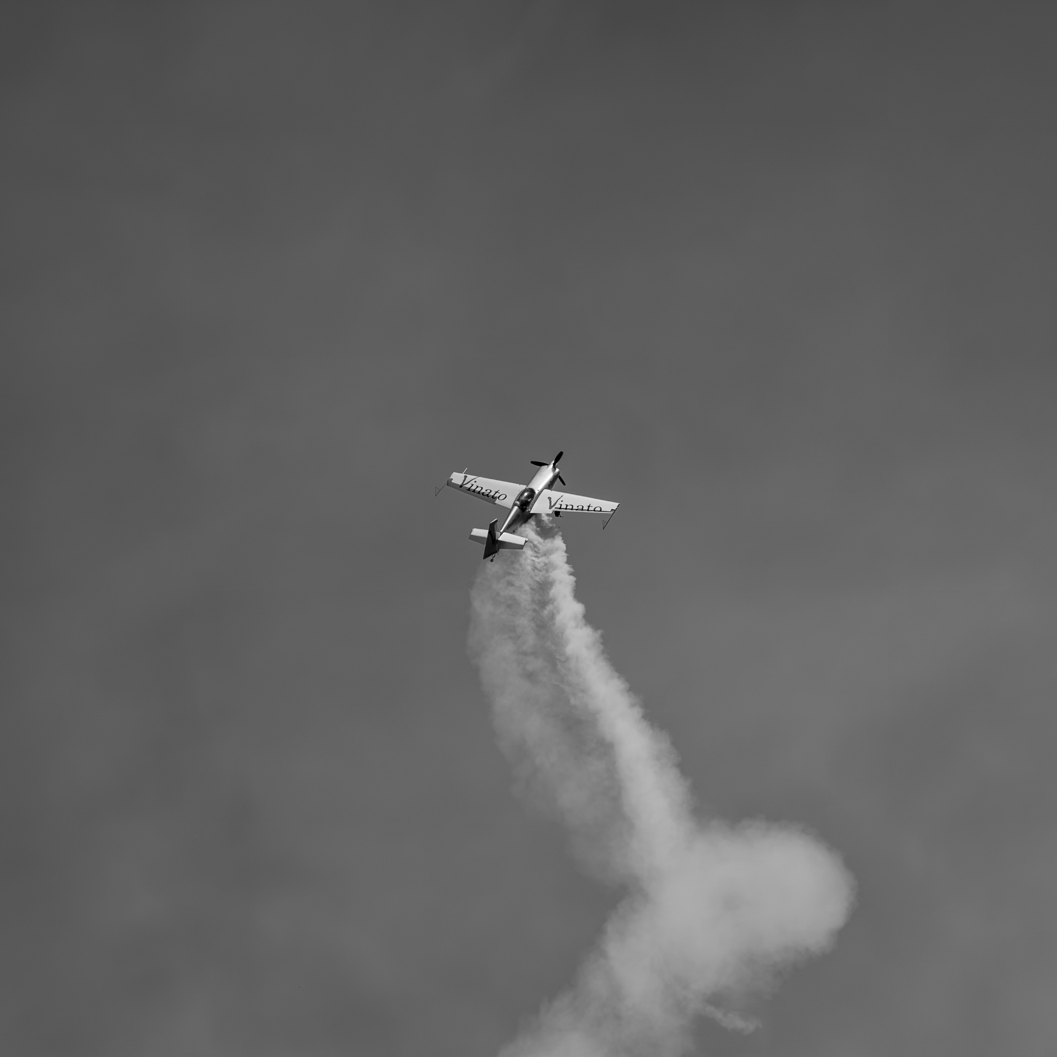 Smoke-trailing aircraft climbs through a vast gray sky, leaving a defined white plume. The image captures a moment of aerial performance with a lone plane against a minimal backdrop, emphasizing motion and scale. (Logo/airshow context).