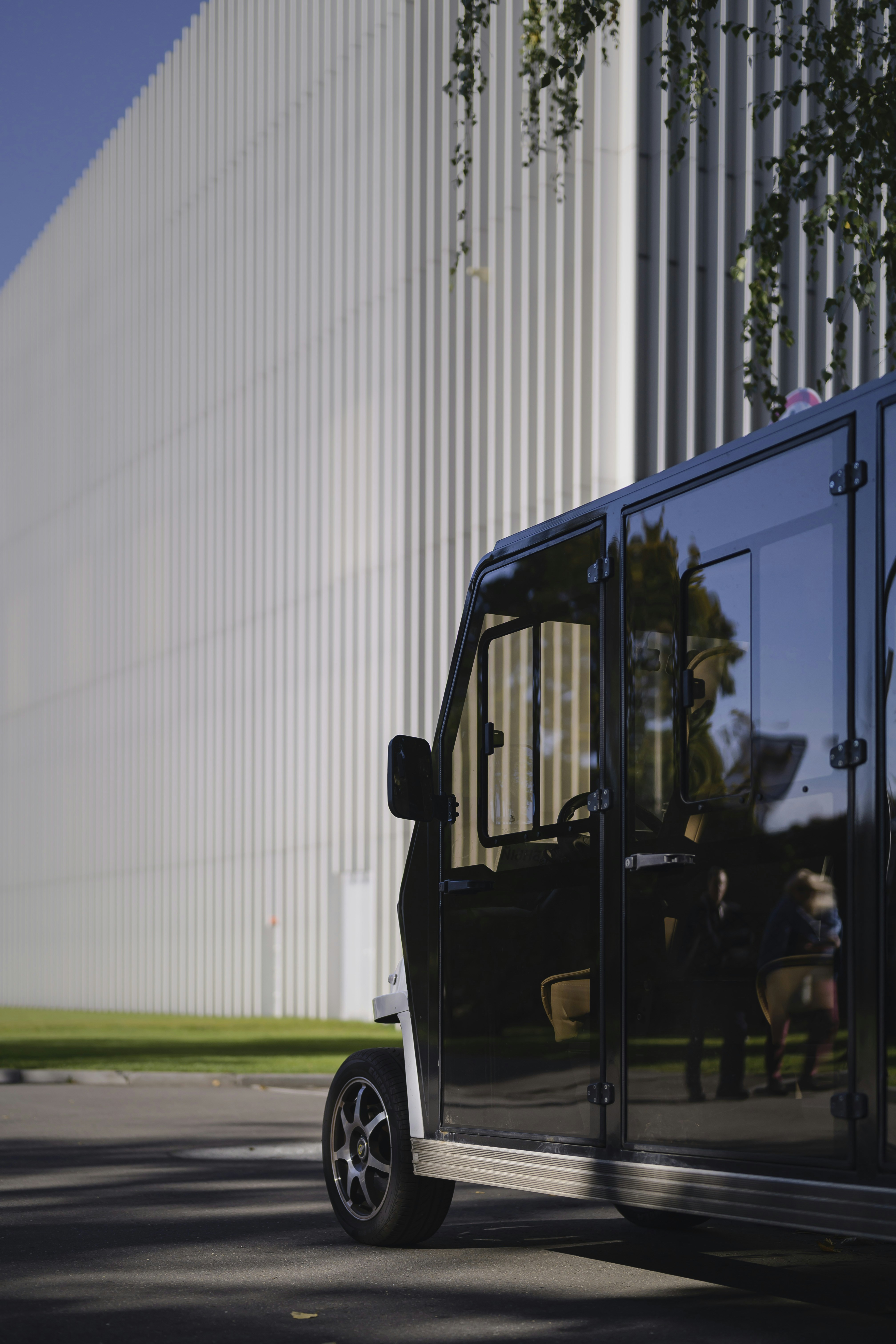 A quiet urban scene where a glossy black utility vehicle parks against a towering façade of vertical metal fins. The image balances clean architectural rhythm and repeating lines with warm reflections, soft shadows, and a hint of overhanging foliage — a study in contrast between human-scale mobility and large‑scale contemporary design.