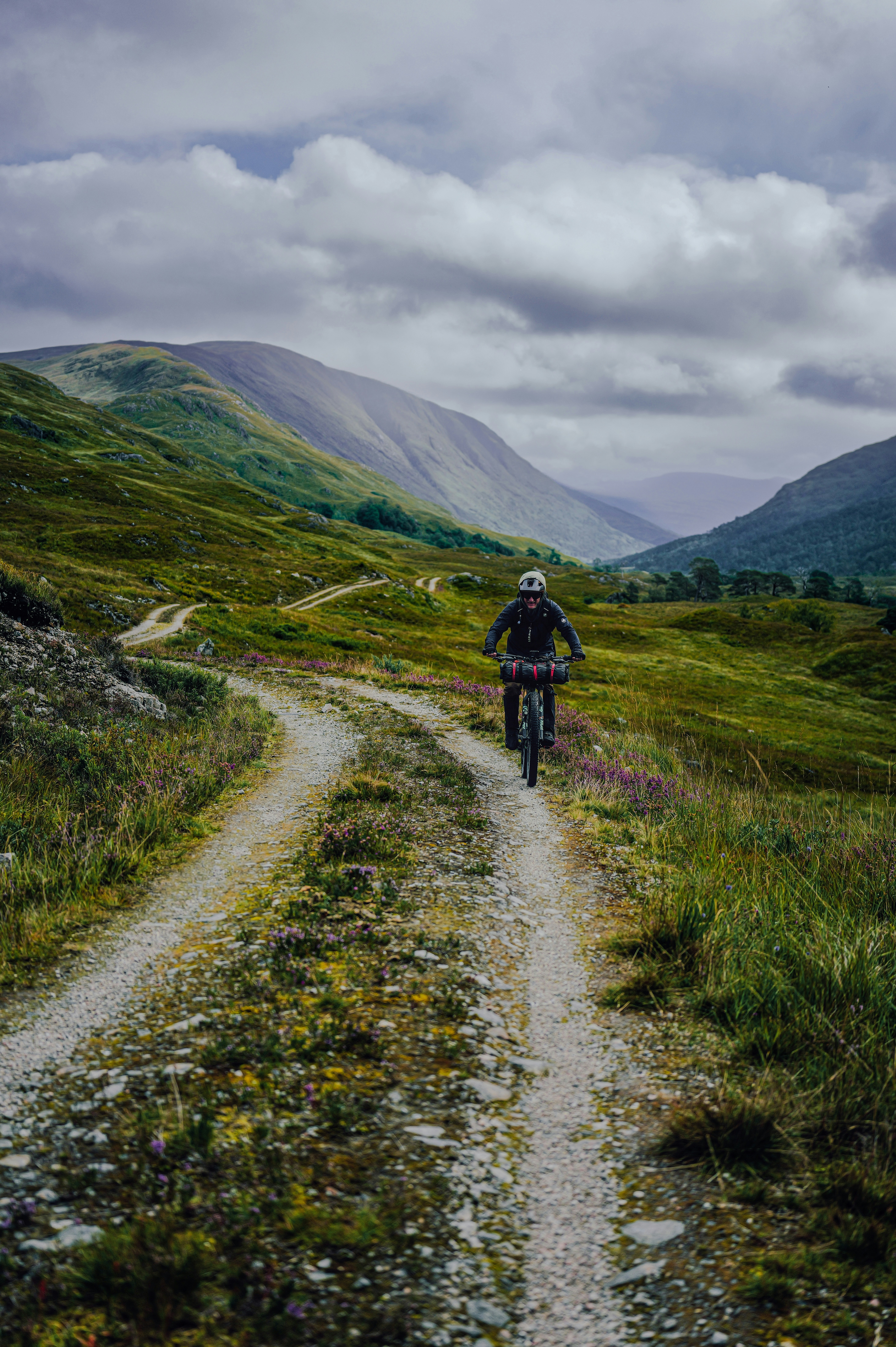 Rugged gravel track winds through rolling Highland hills with a lone cyclist approaching in the distance. A moody sky and vibrant heather add color to the landscape.