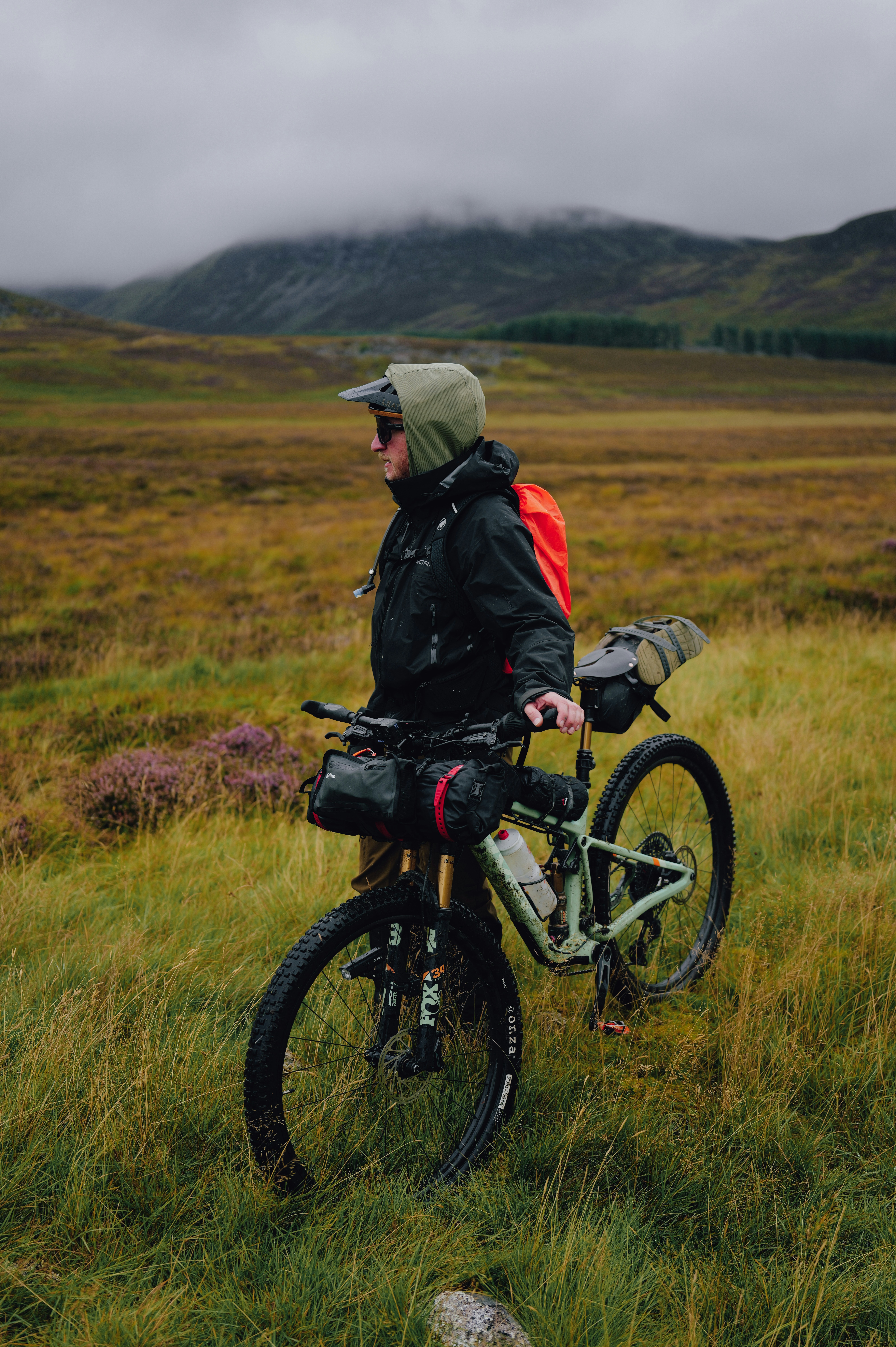 Mountain-biking explorer pauses on a grassy moor with a loaded bike under a moody, clouded sky; gear and trail bags emphasize preparation. This is a photographic capture, not a painting or illustration.