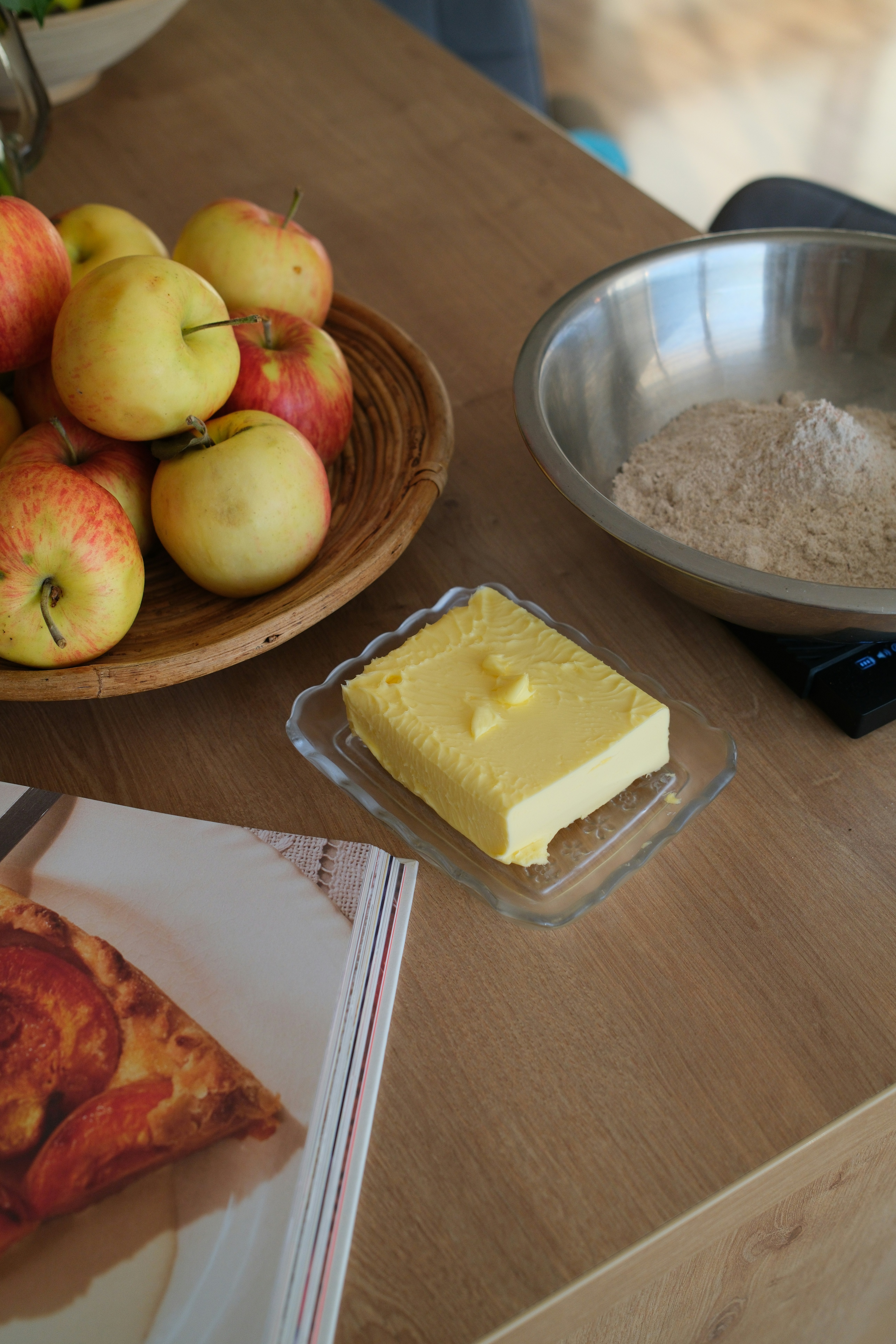 Apples in a wooden basket sit beside a pat of butter on a glass dish and a bowl of flour, with a cookbook partially visible on a wooden counter.