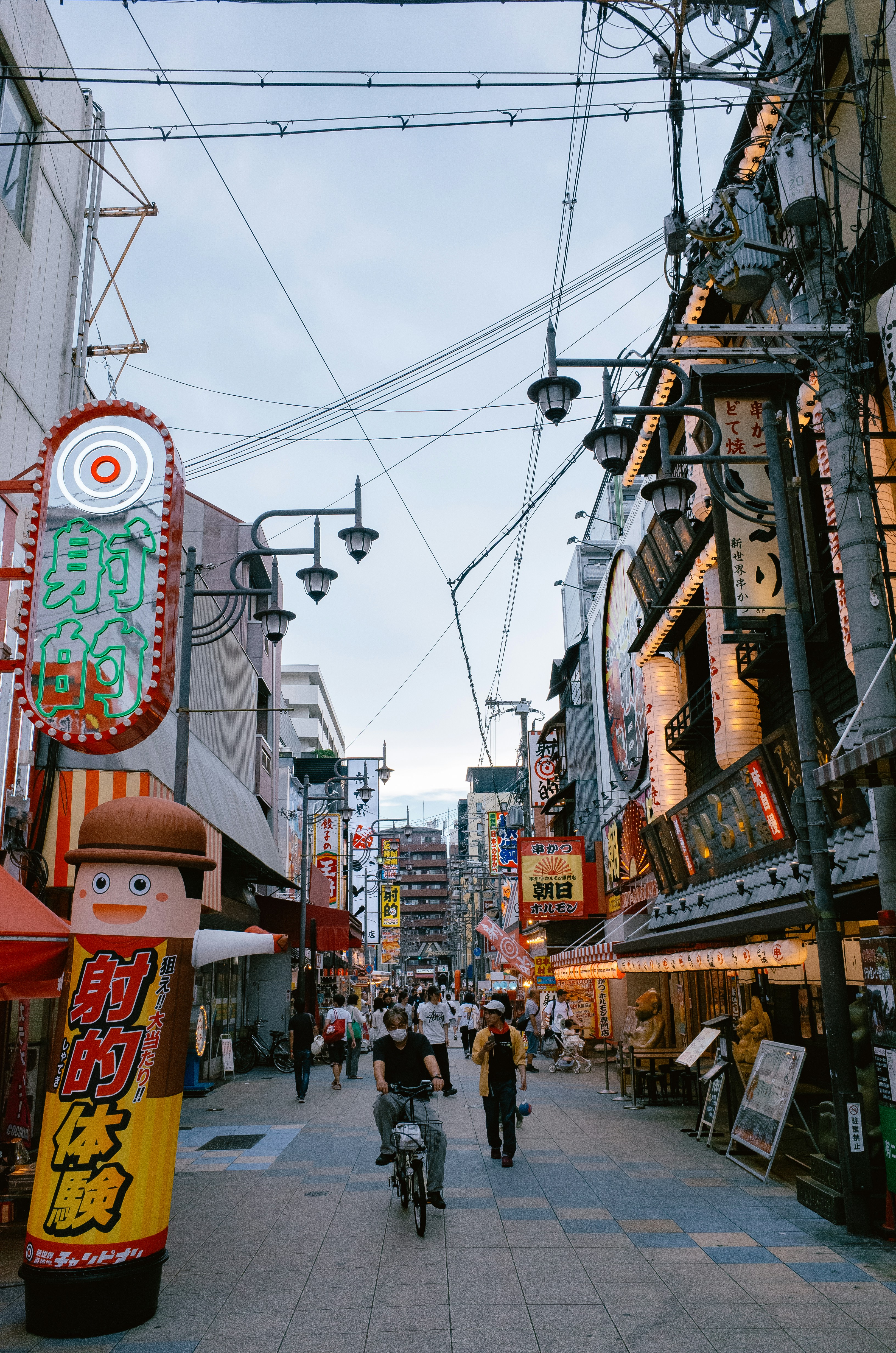 Busy street lined with neon signs and storefronts; pedestrians and a cyclist navigate a vibrant urban corridor with overhead wires. A playful mascot and bold vertical signage give a lively, cinematic feel.