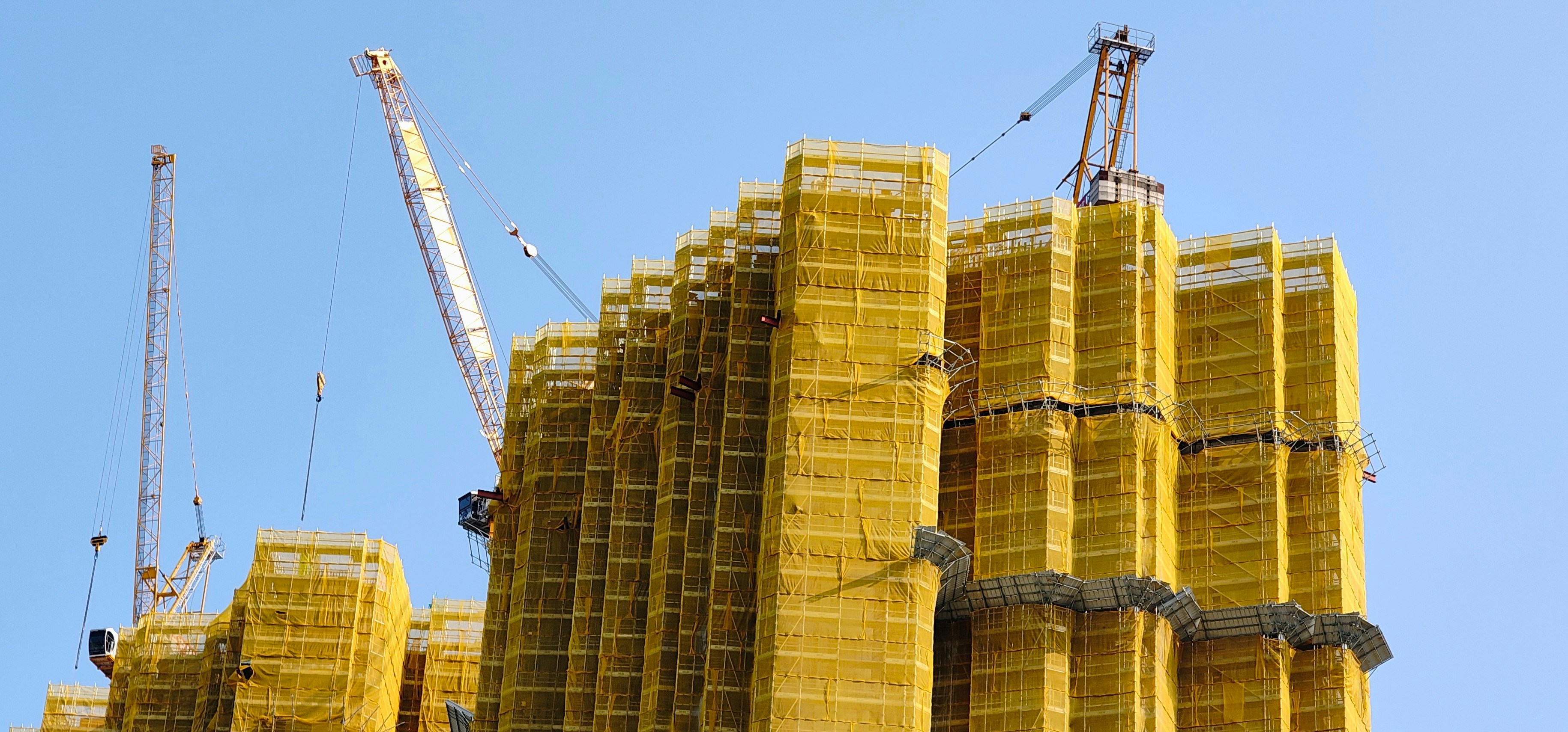 Bright scaffolding towers rise over yellow safety netting under a clear sky