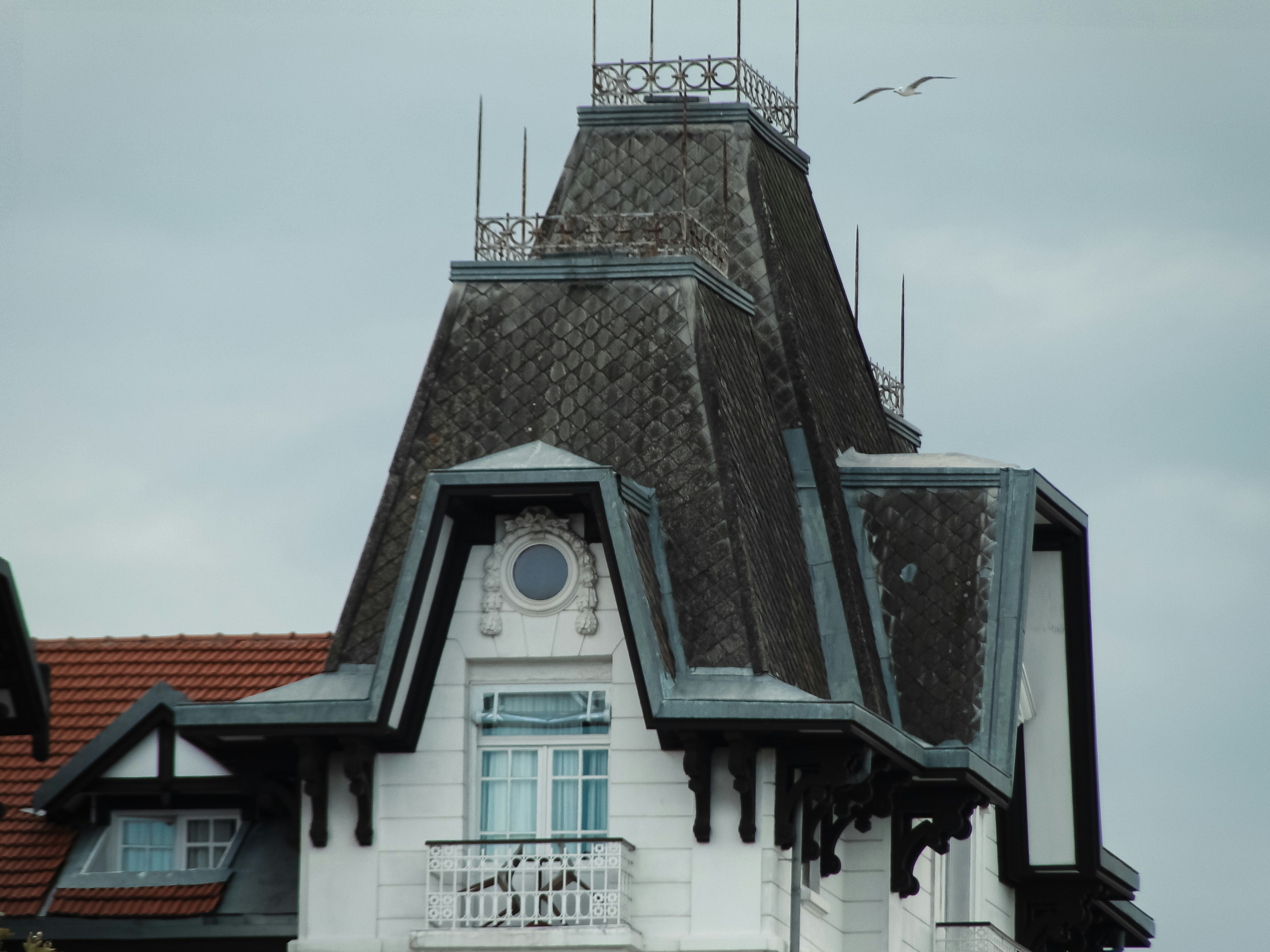 Tudor-style rooftop with steep slate slopes and decorative ironwork, framed by a pale sky and a distant seagull, highlighting architectural detailing.