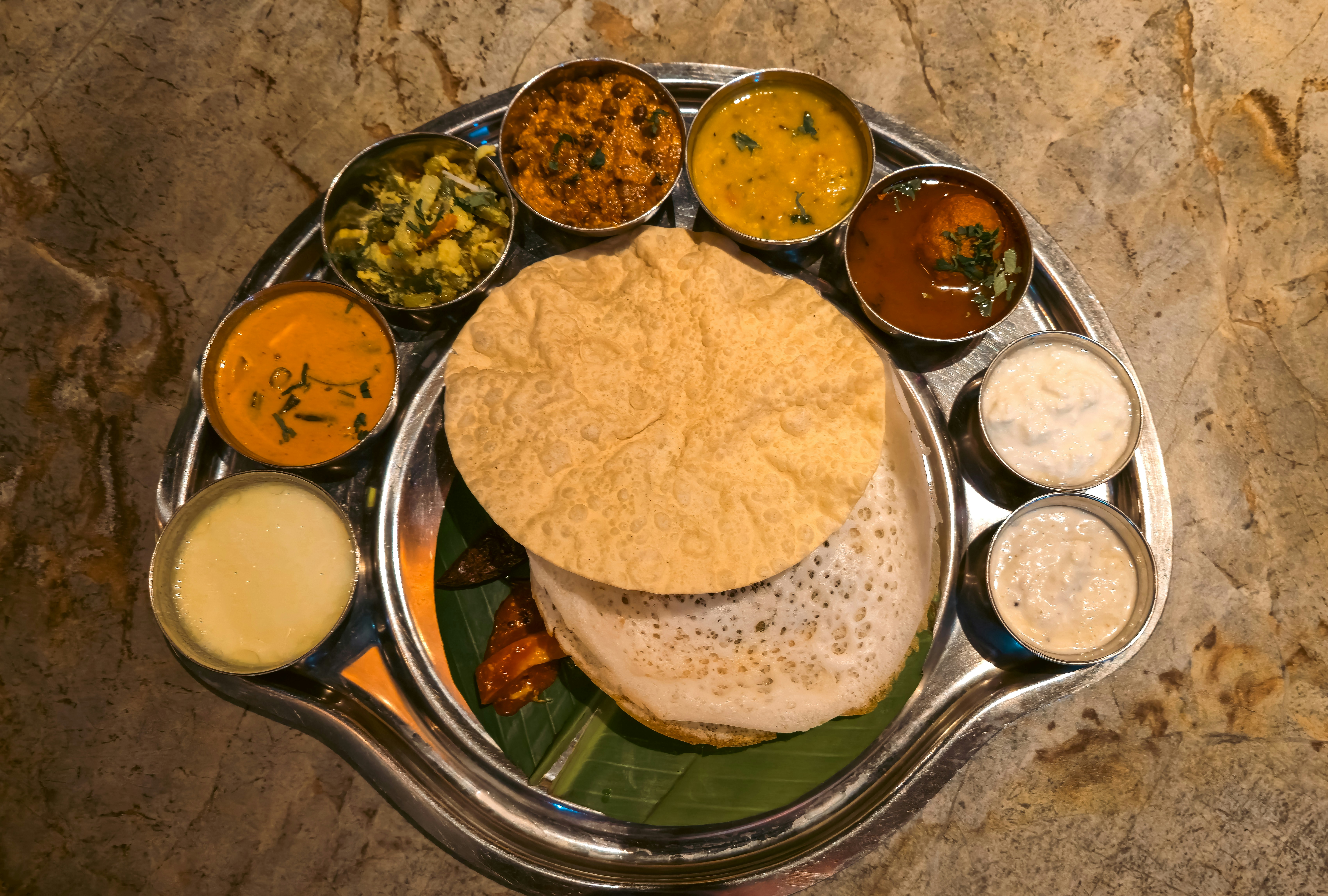 A traditional South Indian thali centered around fluffy, bowl-shaped appams with soft centers and crispy lacy edges, accompanied by an assortment of rich regional curries and side dishes. The vibrant spread highlights the distinct flavors of coastal South Indian and Kerala culinary traditions. Editorial publications, travel guides, cultural food blogs, and regional culinary features can use this image to illustrate authentic regional feasts, traditional banana leaf dining, or South Asian cuisine.