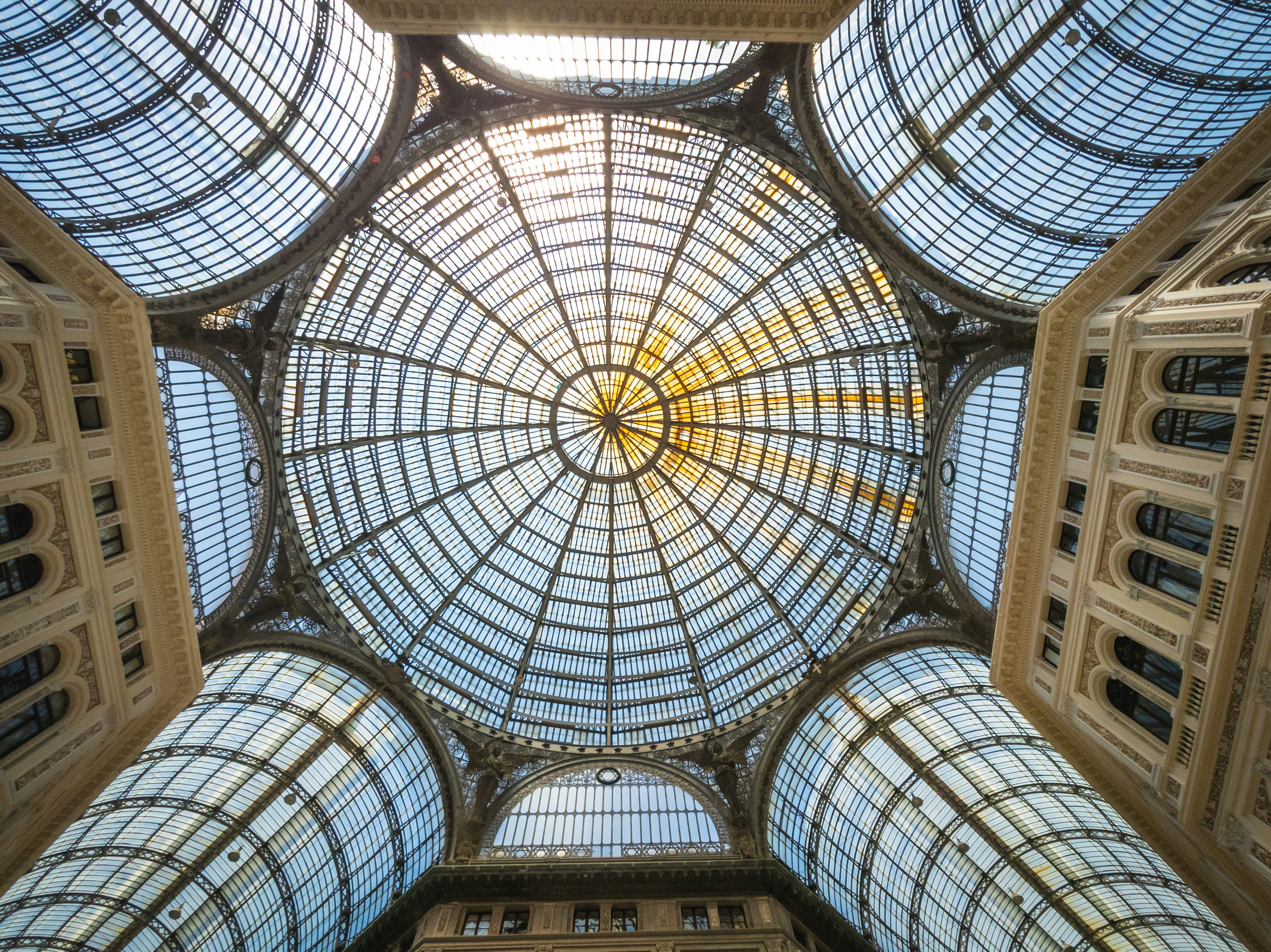 Architectural interior with a vast glass dome ceiling framed by arches and columns, viewed from a low angle to emphasize height and symmetry. This is a photograph of a grand shopping gallery or atrium interior.