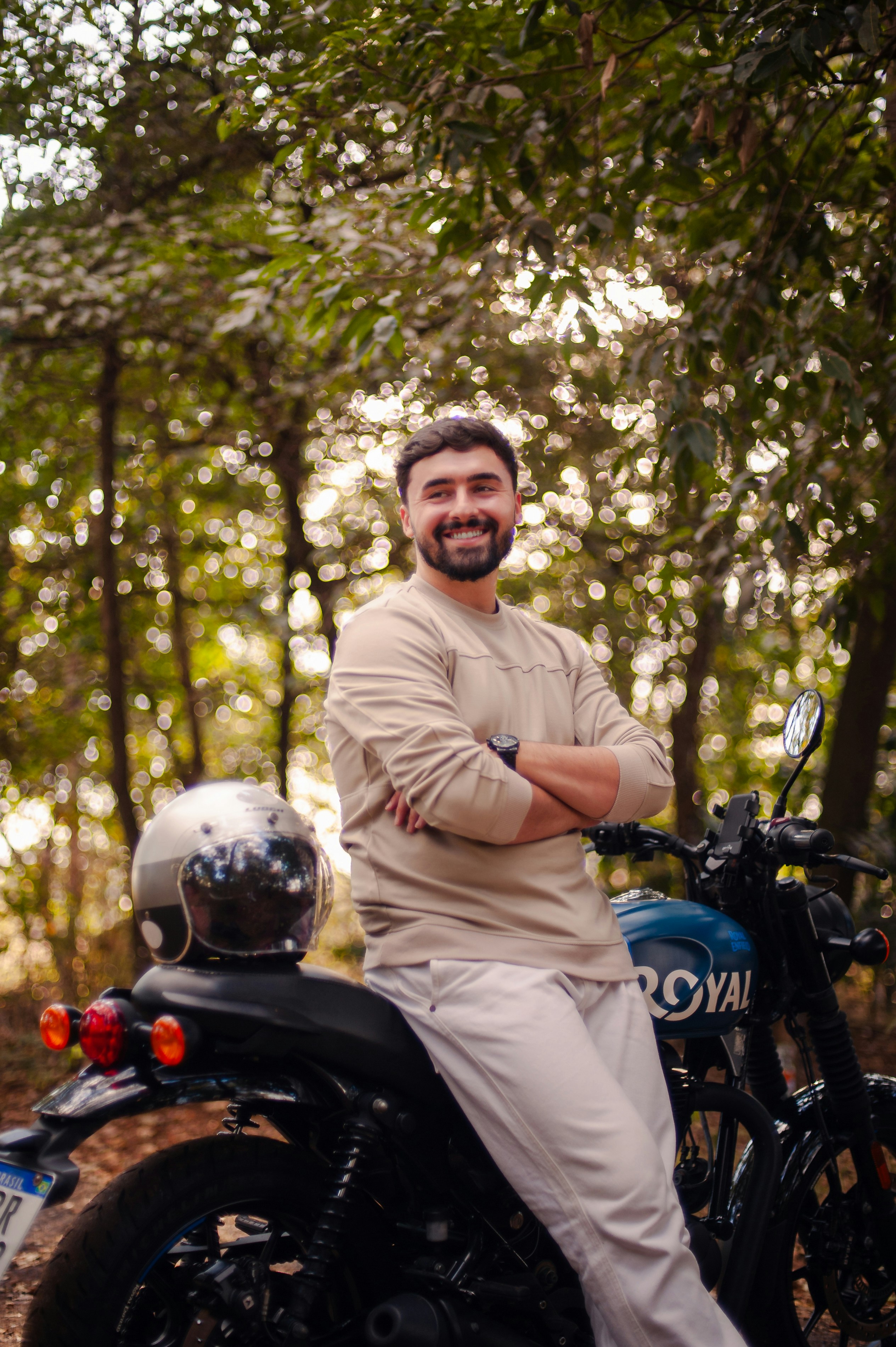 Smiling rider rests on a classic Royal motorcycle in a sun-dappled forest
