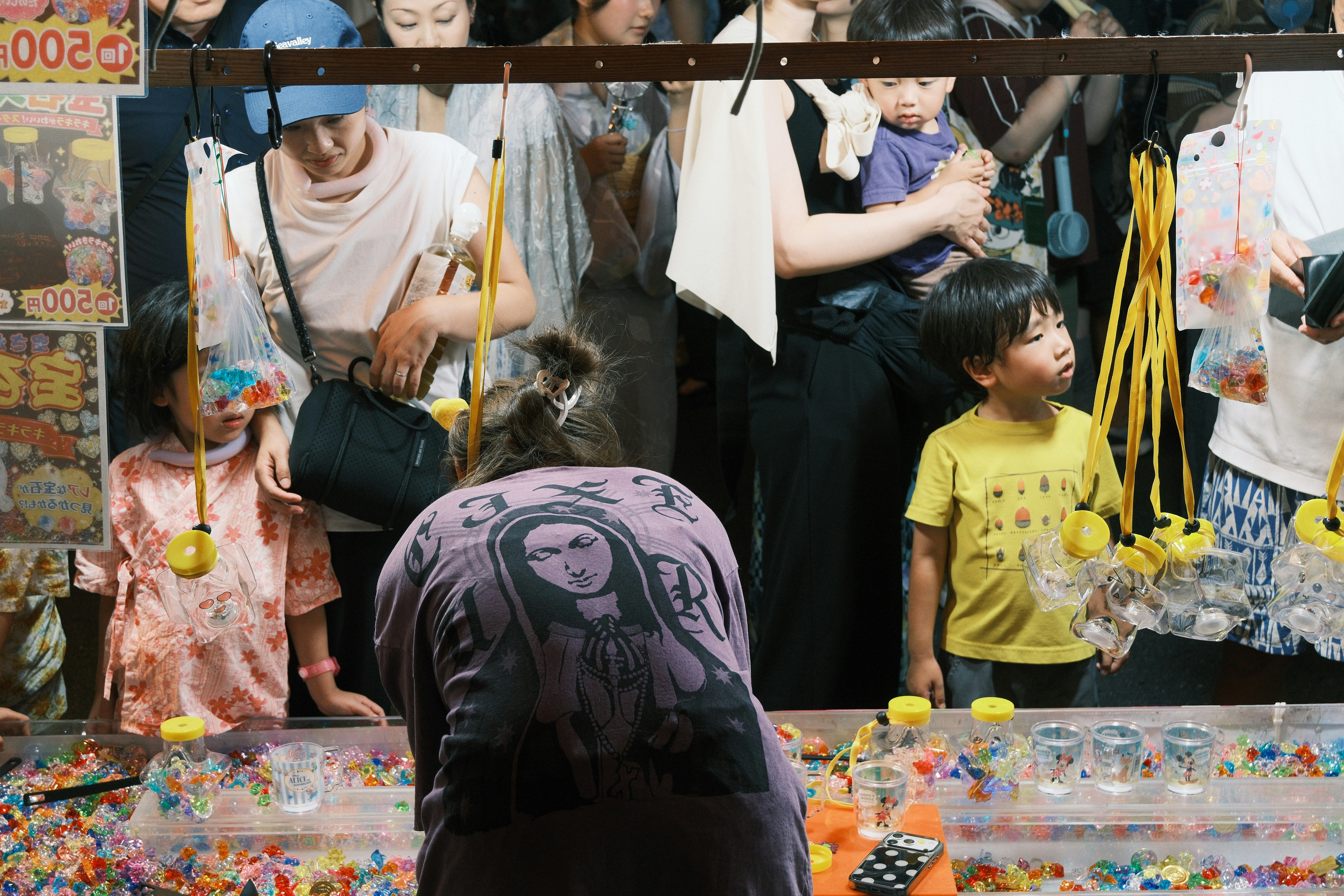 The Crowd Gathers at a Carnival Booth as a Worker Bends Over Colorful Trinkets