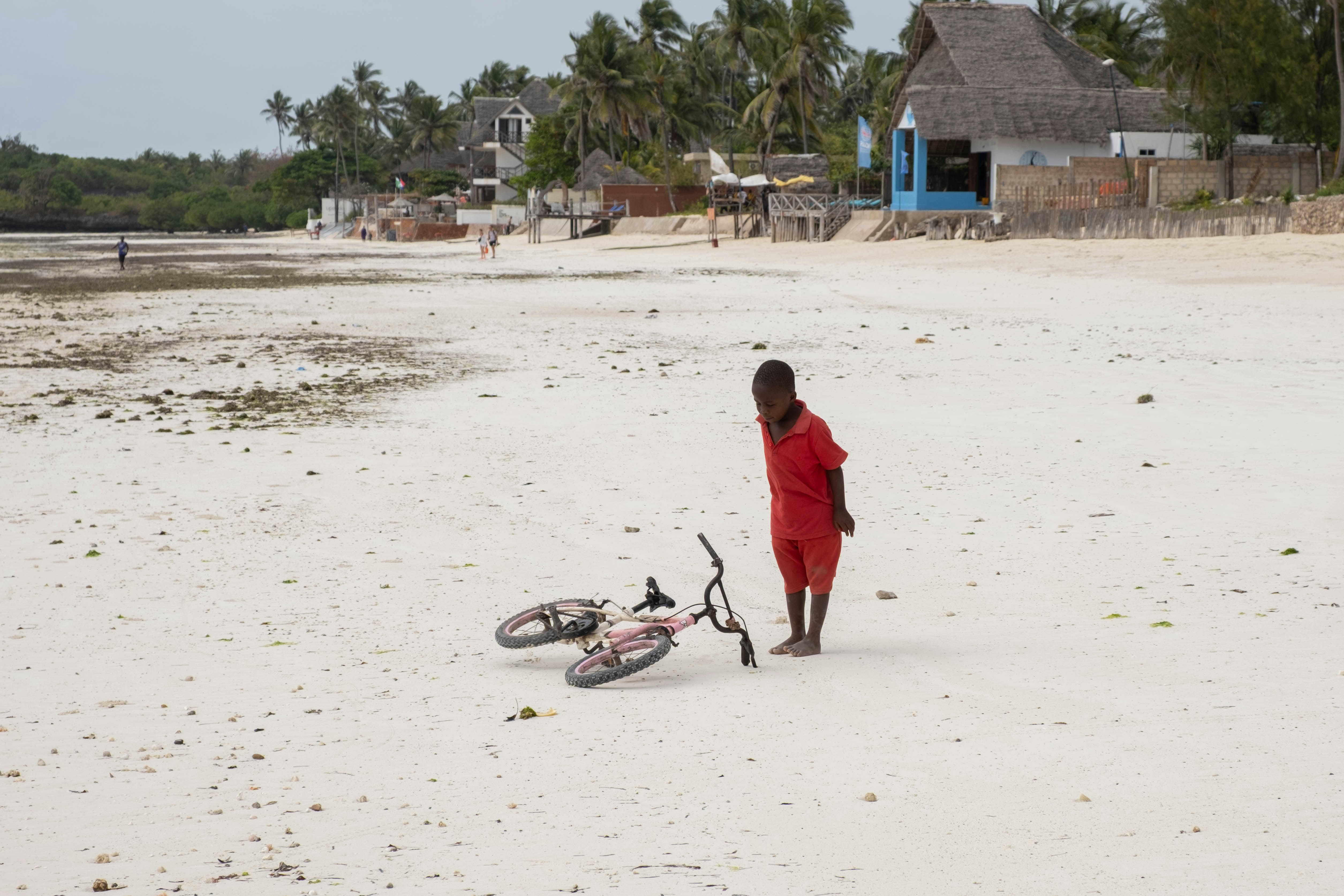 Red-clad child on a sunlit beach beside a fallen bicycle