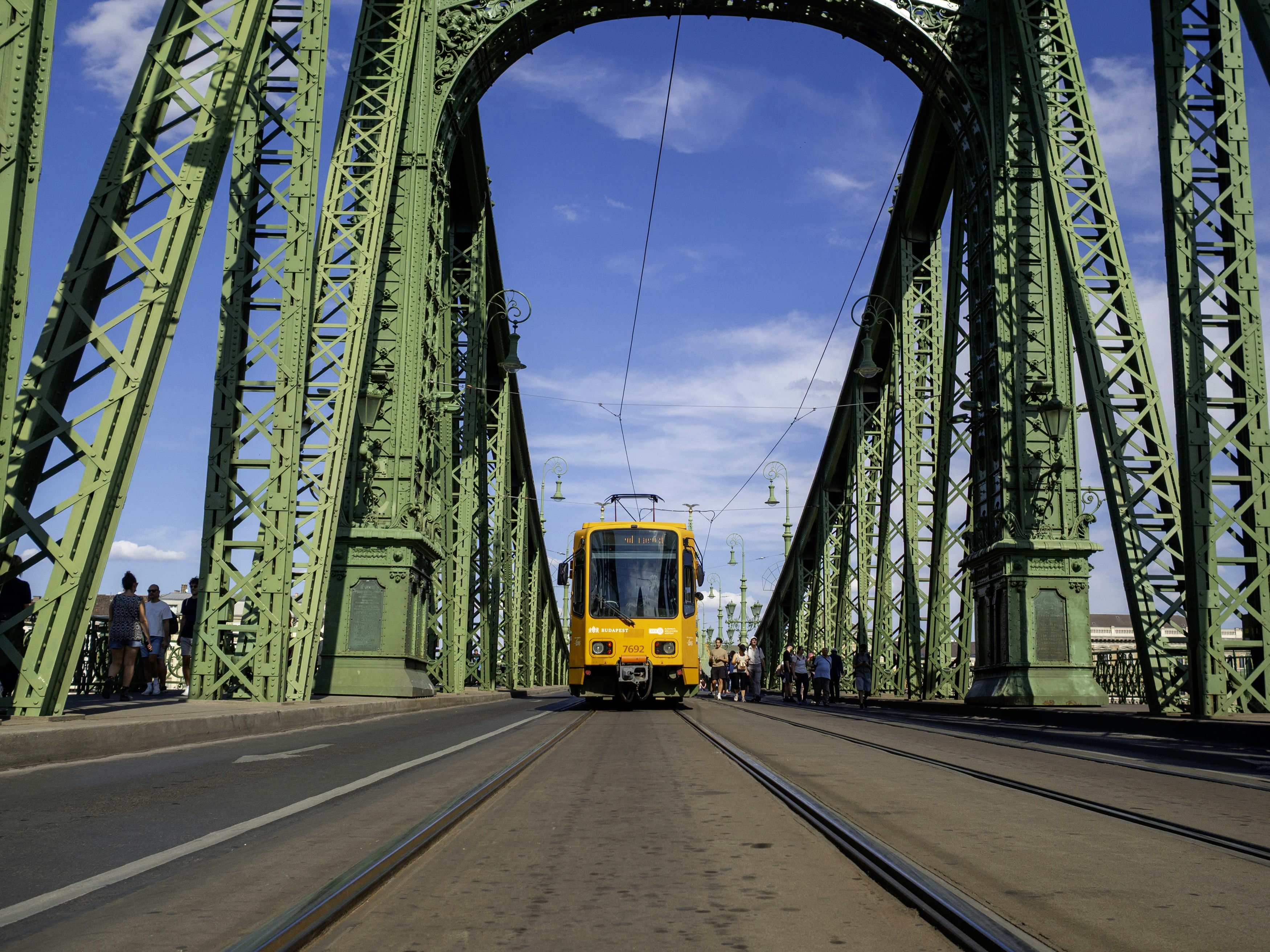 Yellow tram crossing over green steel arches at a sunlit city bridge