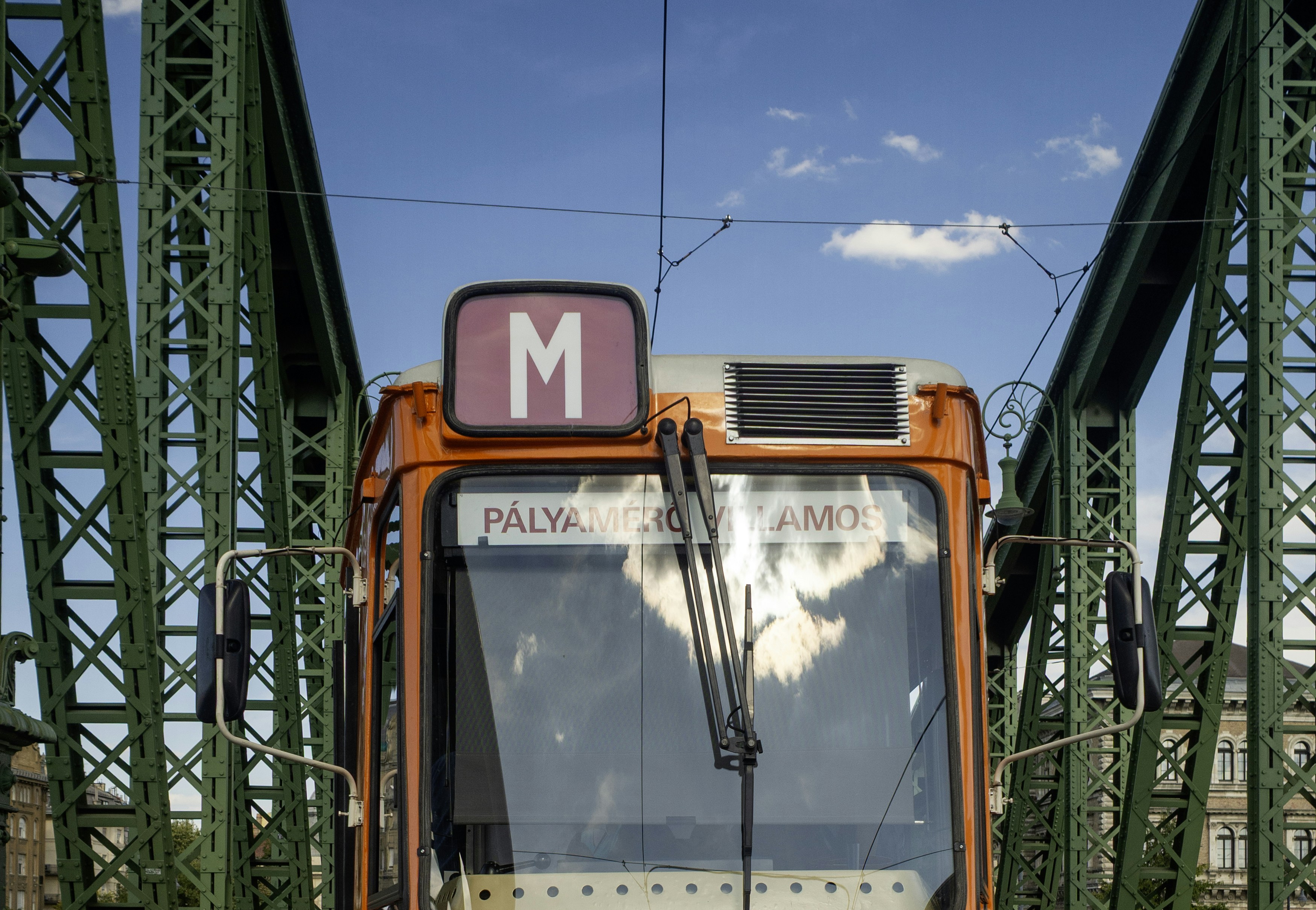 Bright orange tram crosses a green truss bridge, front-facing cab with large windshield and overhead M sign; clear blue sky reflected in glass.