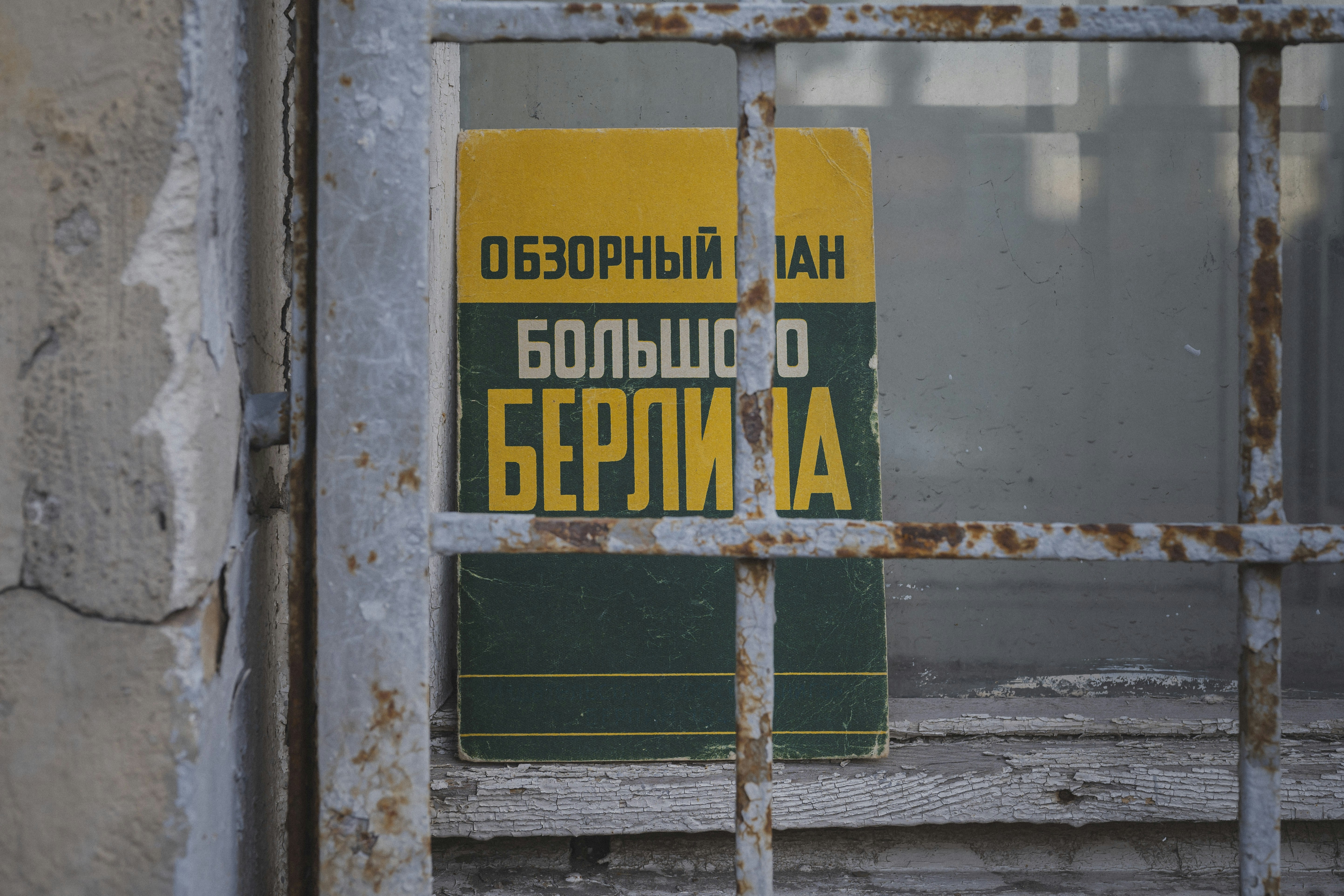 A weathered guidebook to Greater Berlin sits behind a rusty window grille on a cracked wooden sill — its yellow-and-green cover and bold Cyrillic type peeking out from a quiet, forgotten storefront. The composition pairs vintage travel optimism with urban decay: peeling paint, textured metal, and soft, muted light create a nostalgic, cinematic study of place, memory, and small found objects.