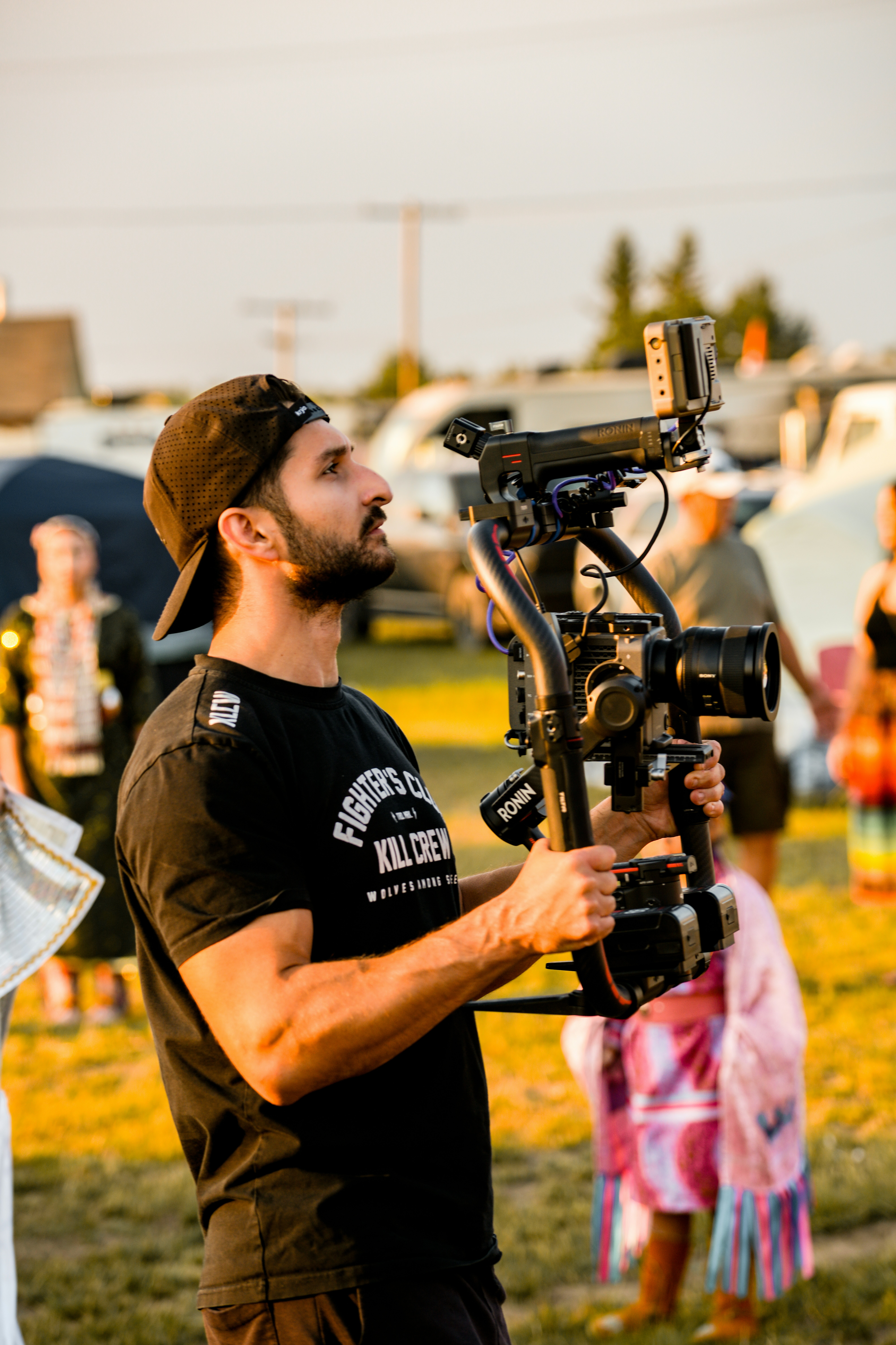 Male videographer in a black t-shirt and backward cap operates a professional rig at an outdoor event, with warm, golden-hour light and blurred background elements.