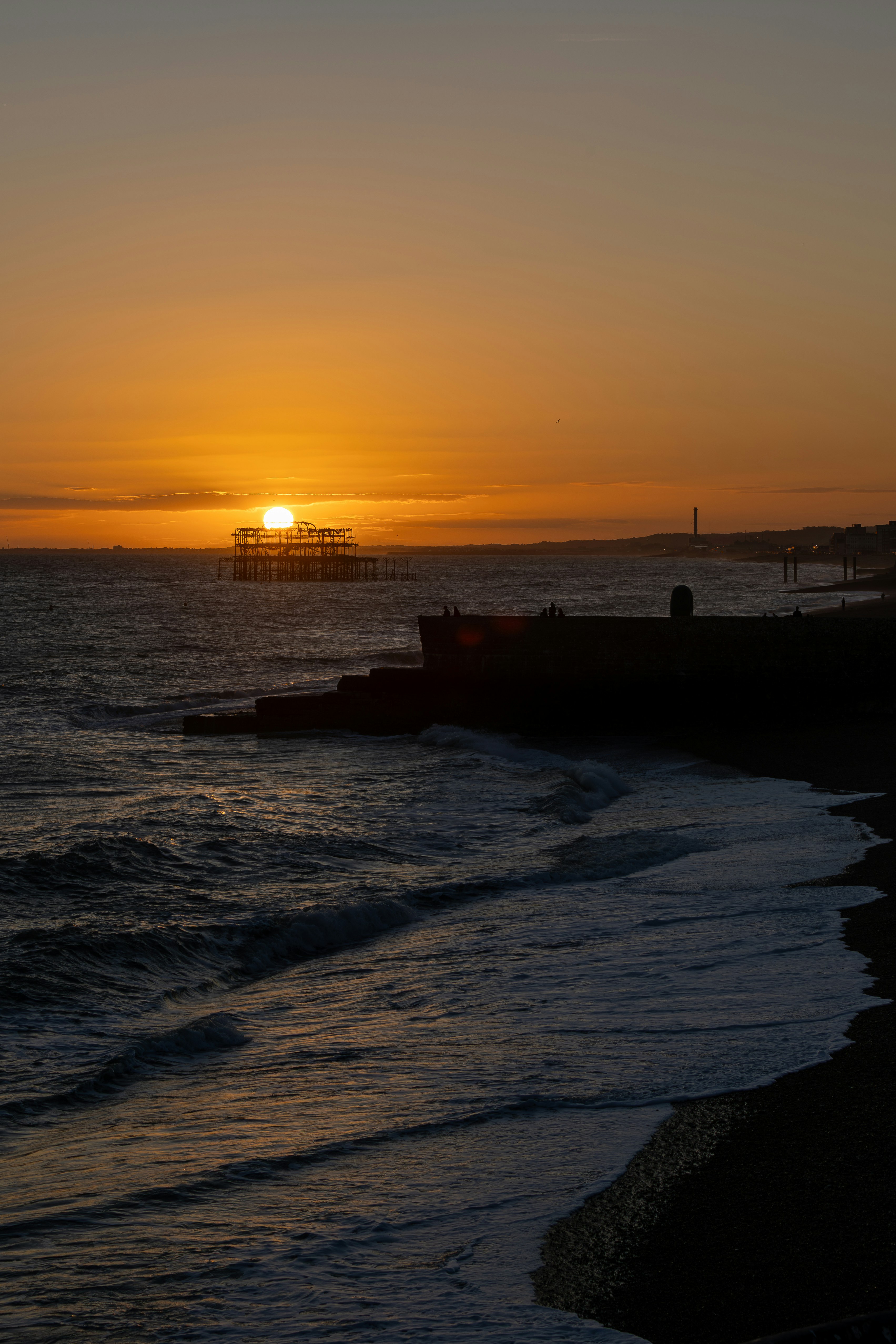 Sunset over a calm sea with a weathered pier silhouette and gentle waves along a dark shoreline.