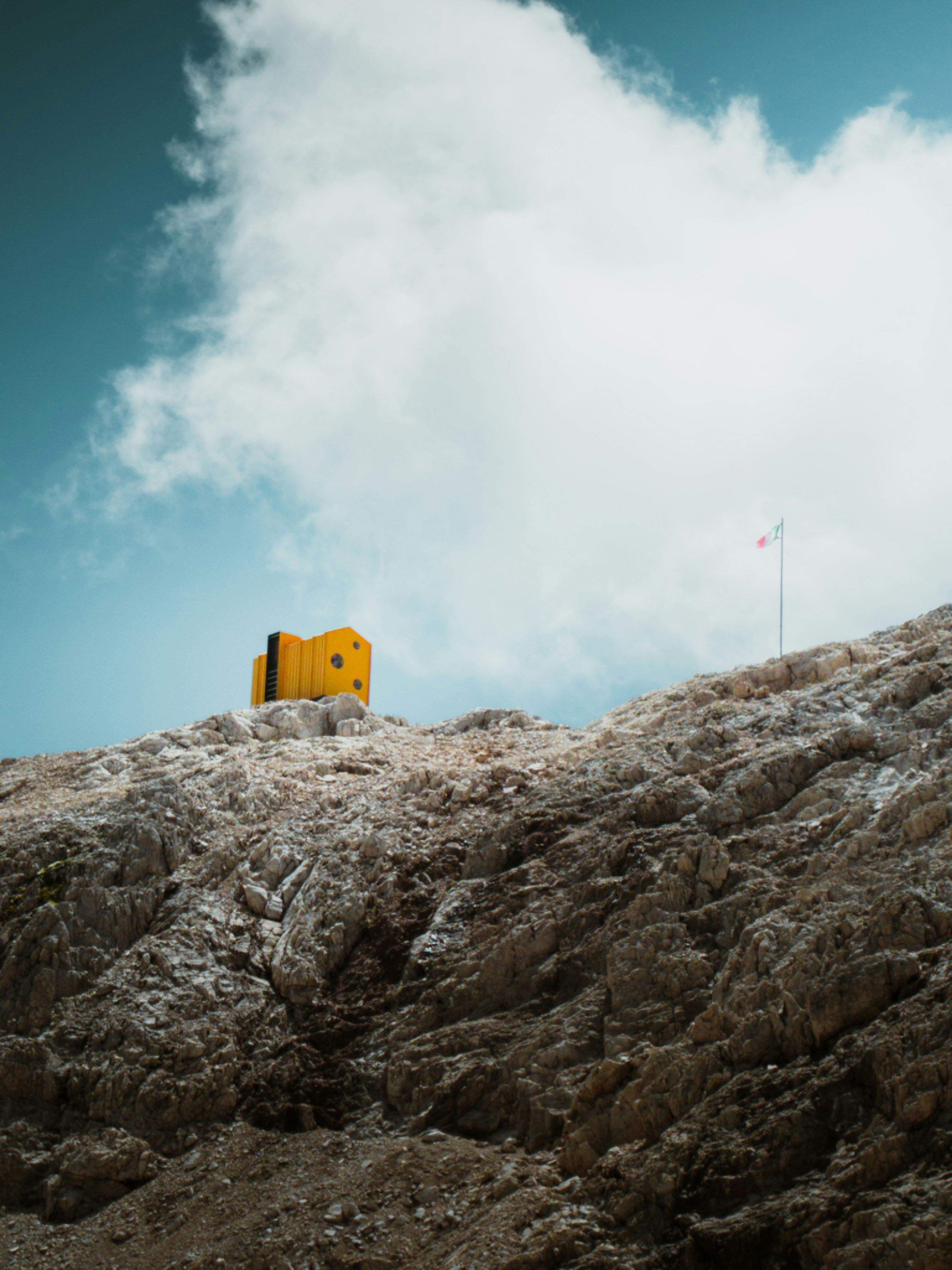 Bright yellow rugged equipment sits atop a rocky outcrop under a blue sky with white clouds. A small flag on a pole adds a subtle human element to the barren terrain.