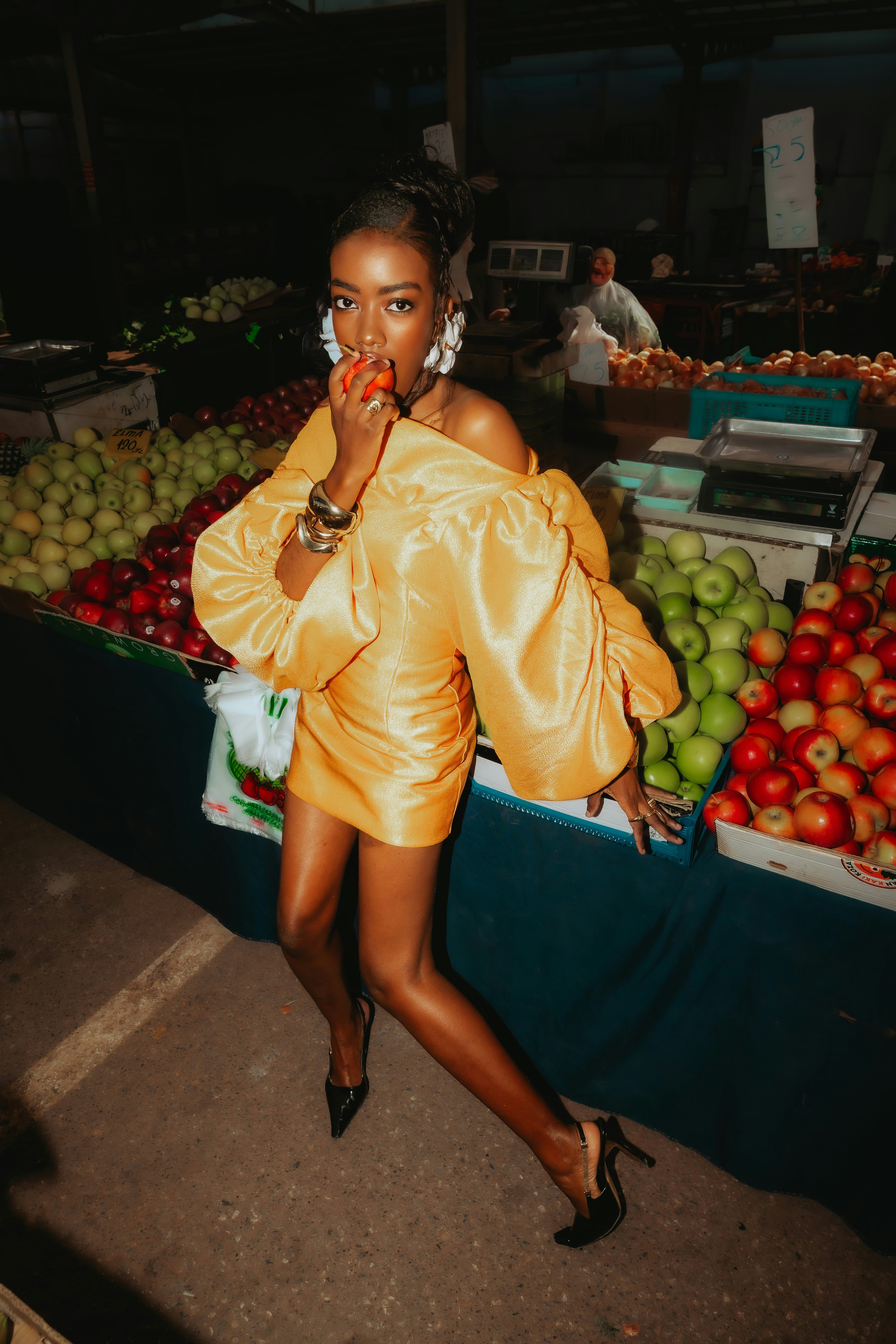 A stylish woman in a bright orange off-shoulder dress poses at a bustling fruit market, holding a fruit close to her lips as she leans against a produce stand. Note: the scene includes a variety of fruits and market activity in the background.