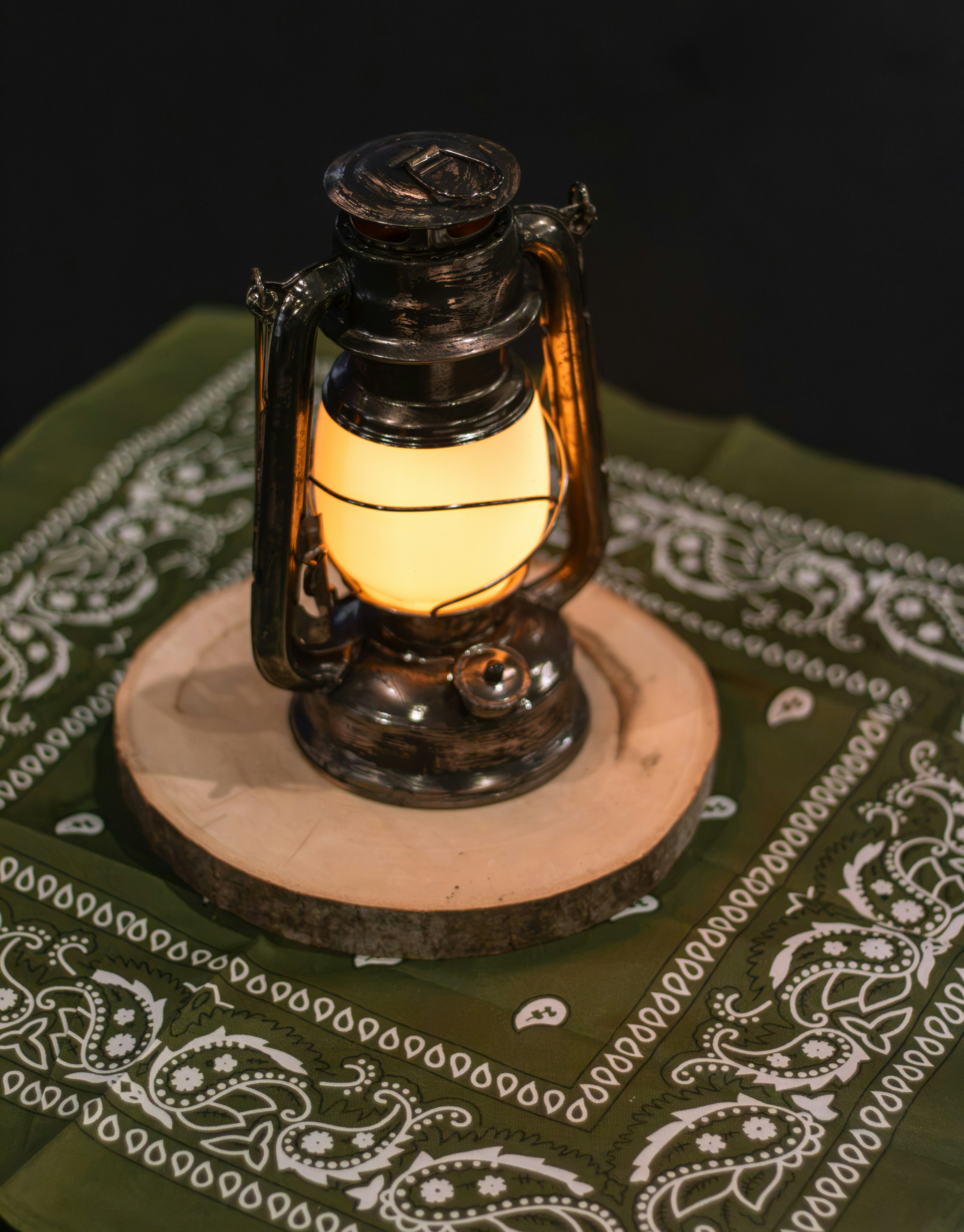 A vintage lantern sits on a wooden slice atop a green bandana, softly illuminated against a dark backdrop.