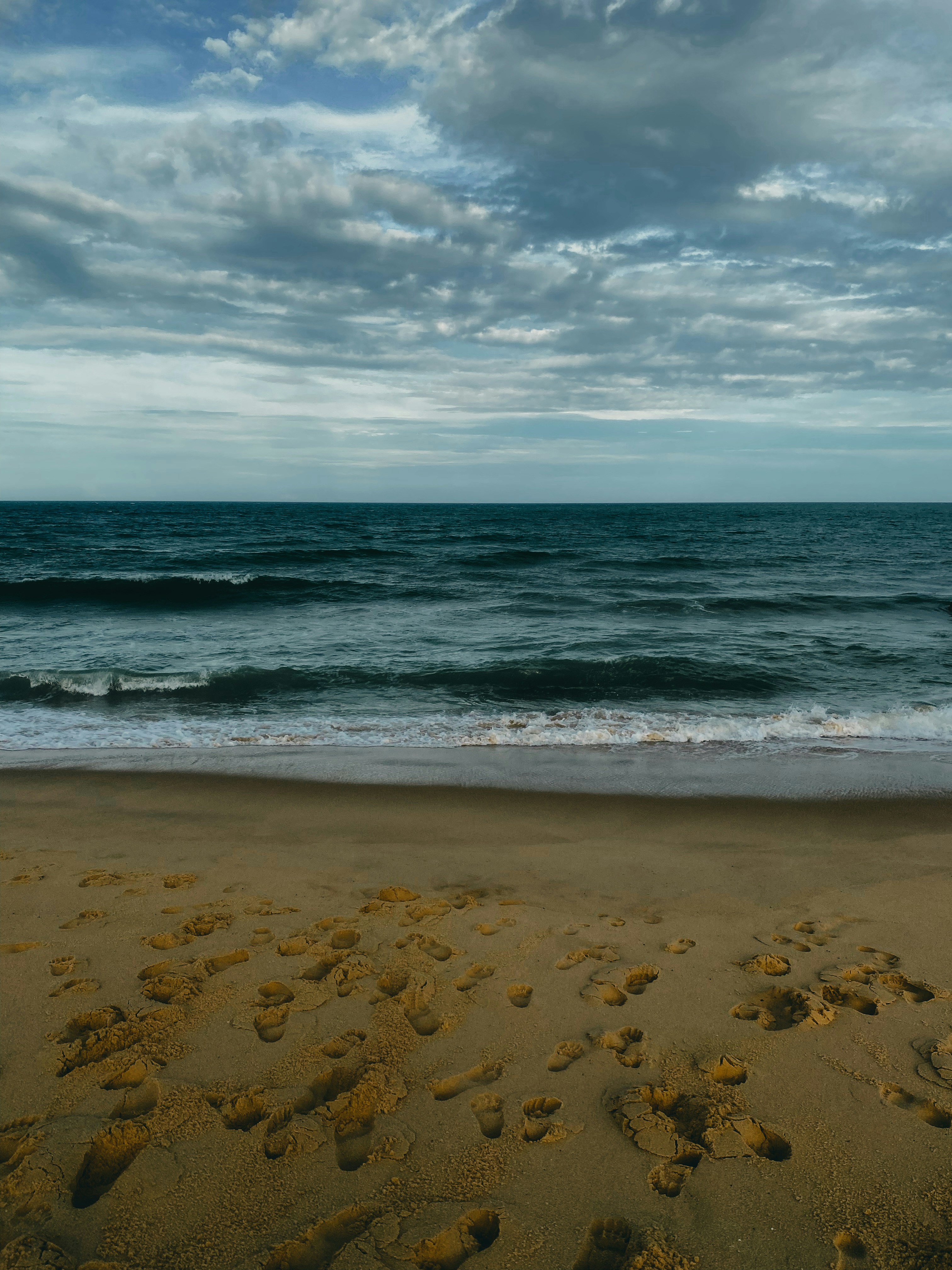 Golden sand in the foreground with scattered footprints, leading to calm waves under a cloudy sky at the shore.