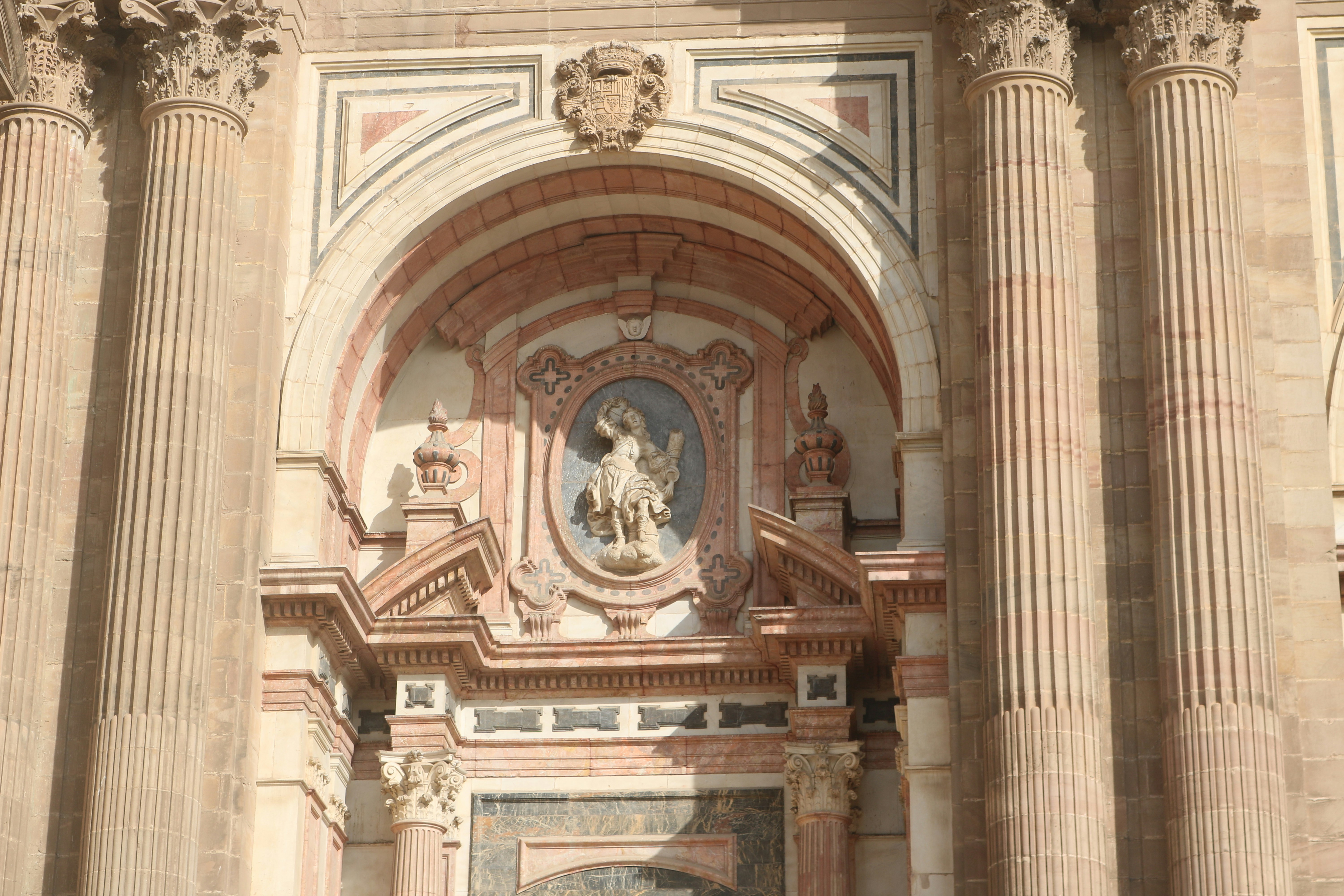 Ornate stone archway with sculpted relief at center, flanked by tall fluted columns; warm daylight highlights architectural detailing and frescoed inner vaults.