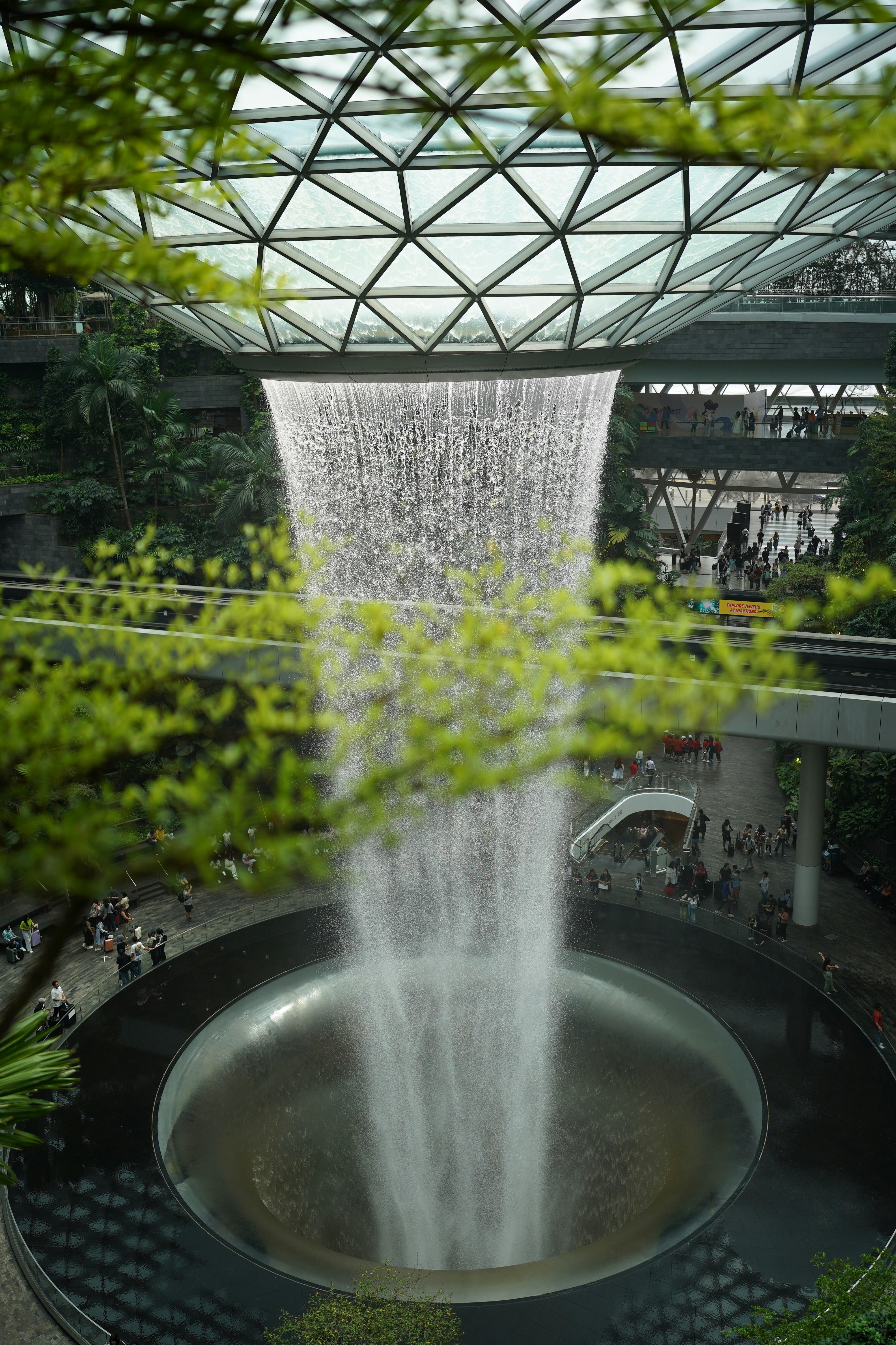 The HSBC Rain Vortex, the indoor waterfall at the centre of Jewel Changi Airport, dropping through the oculus in the glass roof while visitors watch from the levels around it. Singapore.