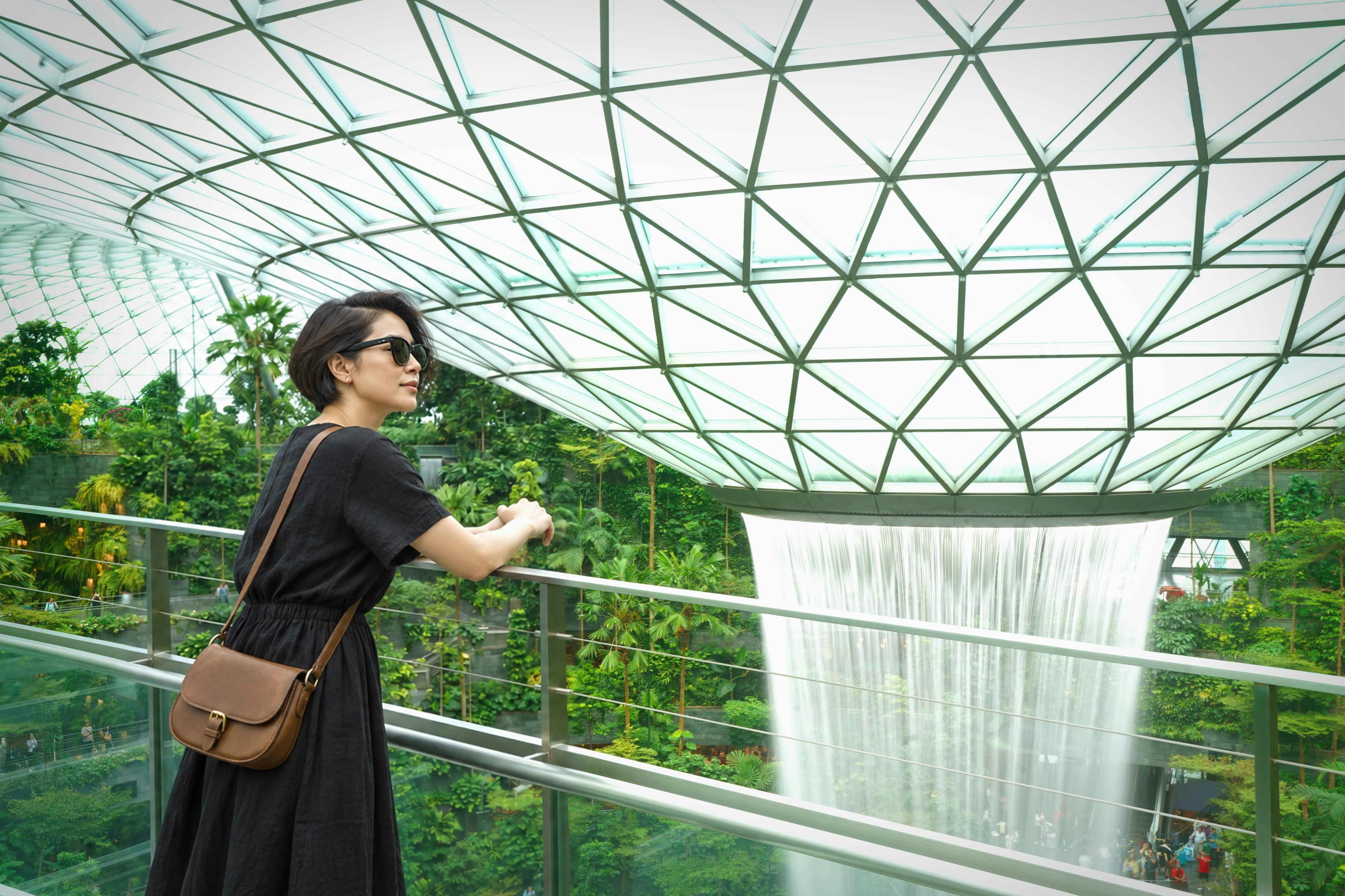 Asian woman poses next to the HSBC Rain Vortex, the indoor waterfall at the centre of Jewel Changi Airport, dropping through the oculus in the glass roof while visitors watch from the levels around it. Singapore.