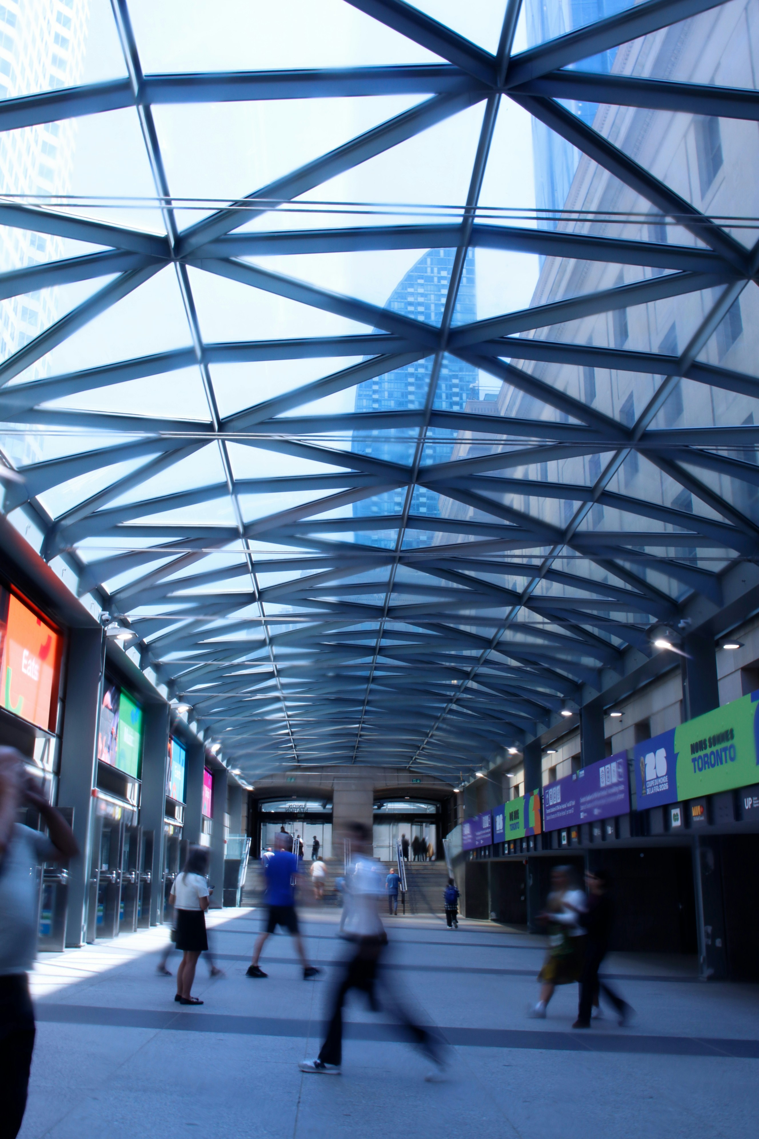 Busy urban transit hall with a geometric glass ceiling and blurred pedestrians; a modern rail-like concourse with signs and platforms visible in perspective.