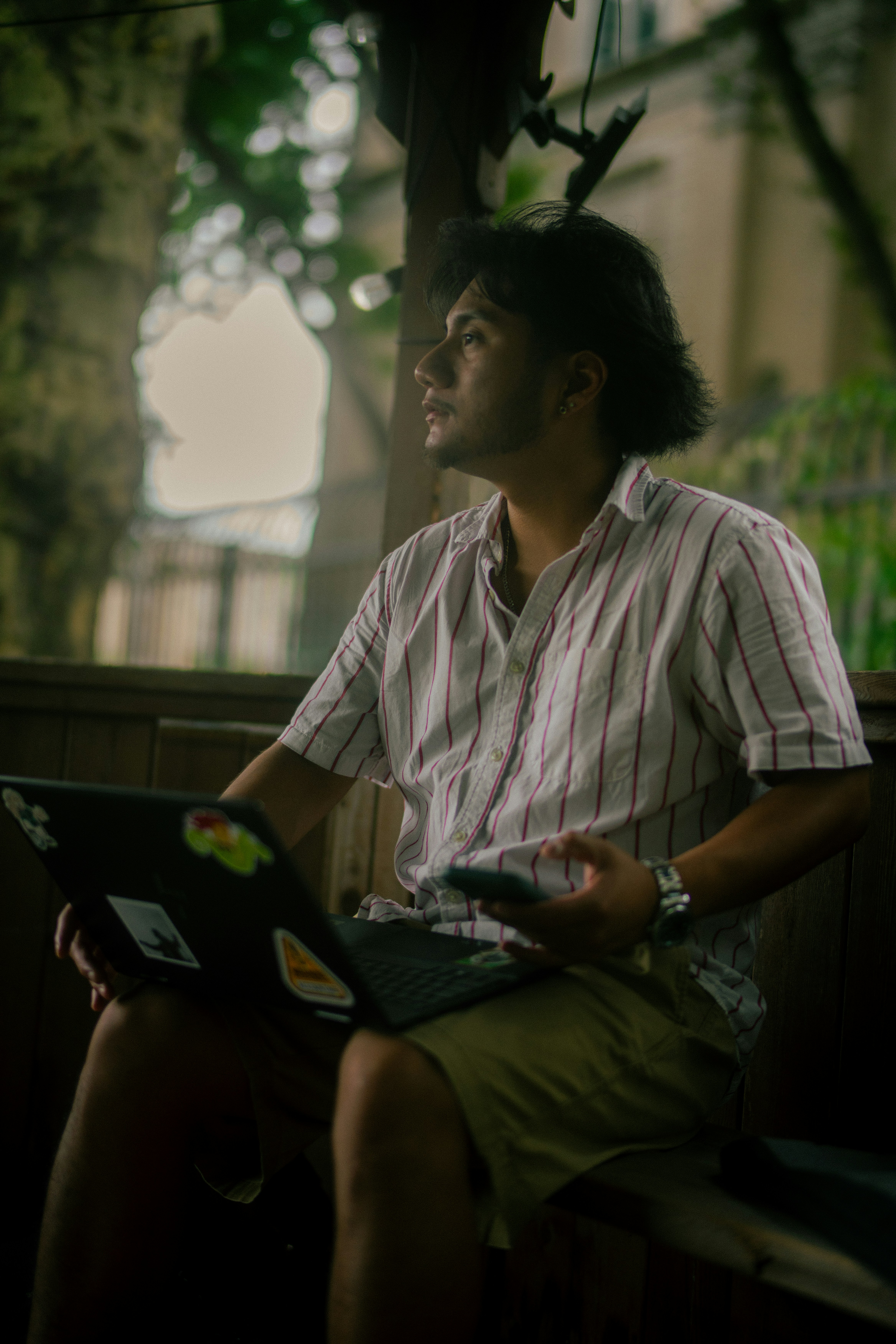 Man seated on a wooden bench with a laptop and phone, bathed in muted outdoor light; the scene emphasizes concentration and a casual working moment. This is a candid portrait with a laptop stickered and a relaxed vibe.