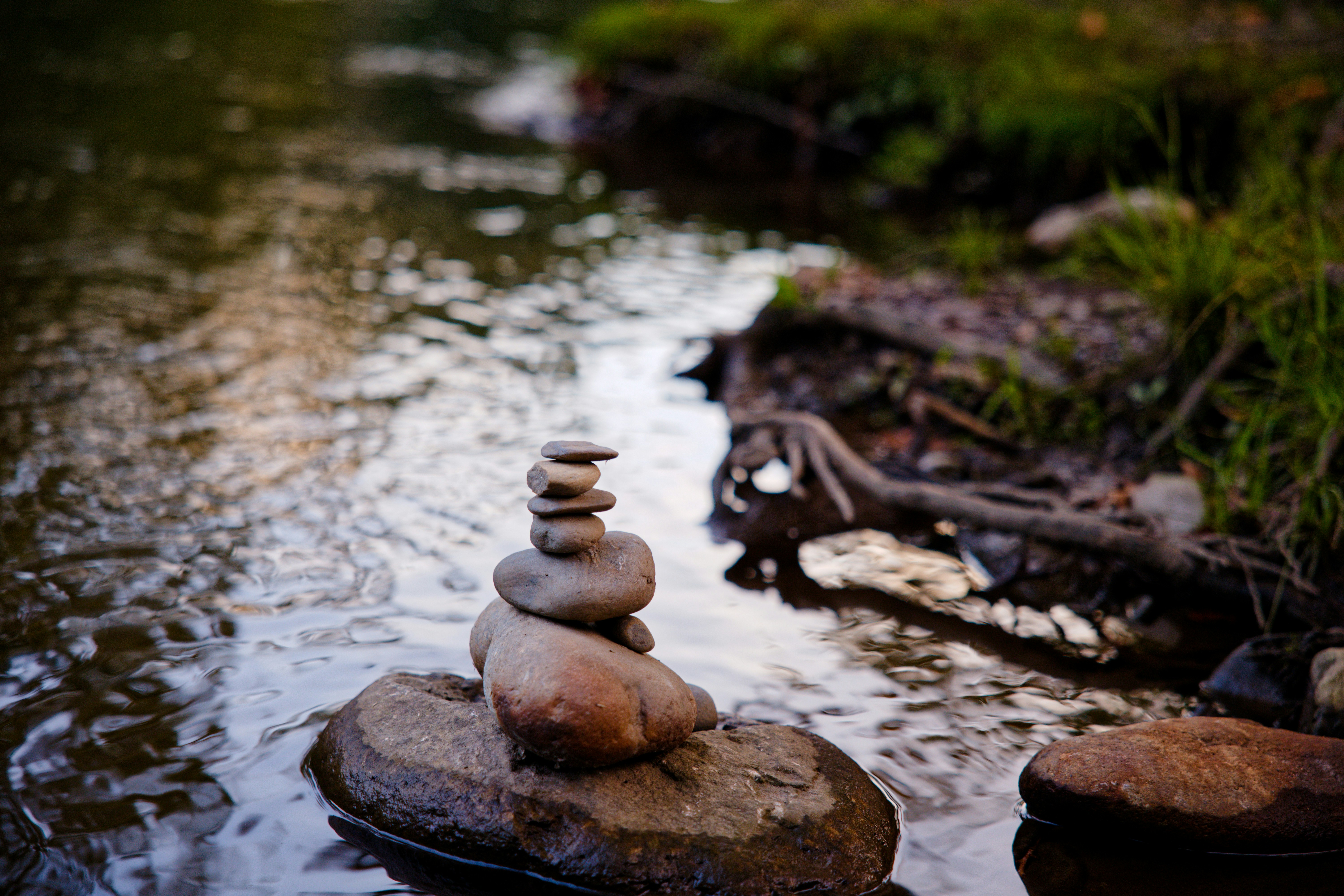 Stacked rocks balanced near a tranquil riverside, with reflective water and surrounding stones and foliage.