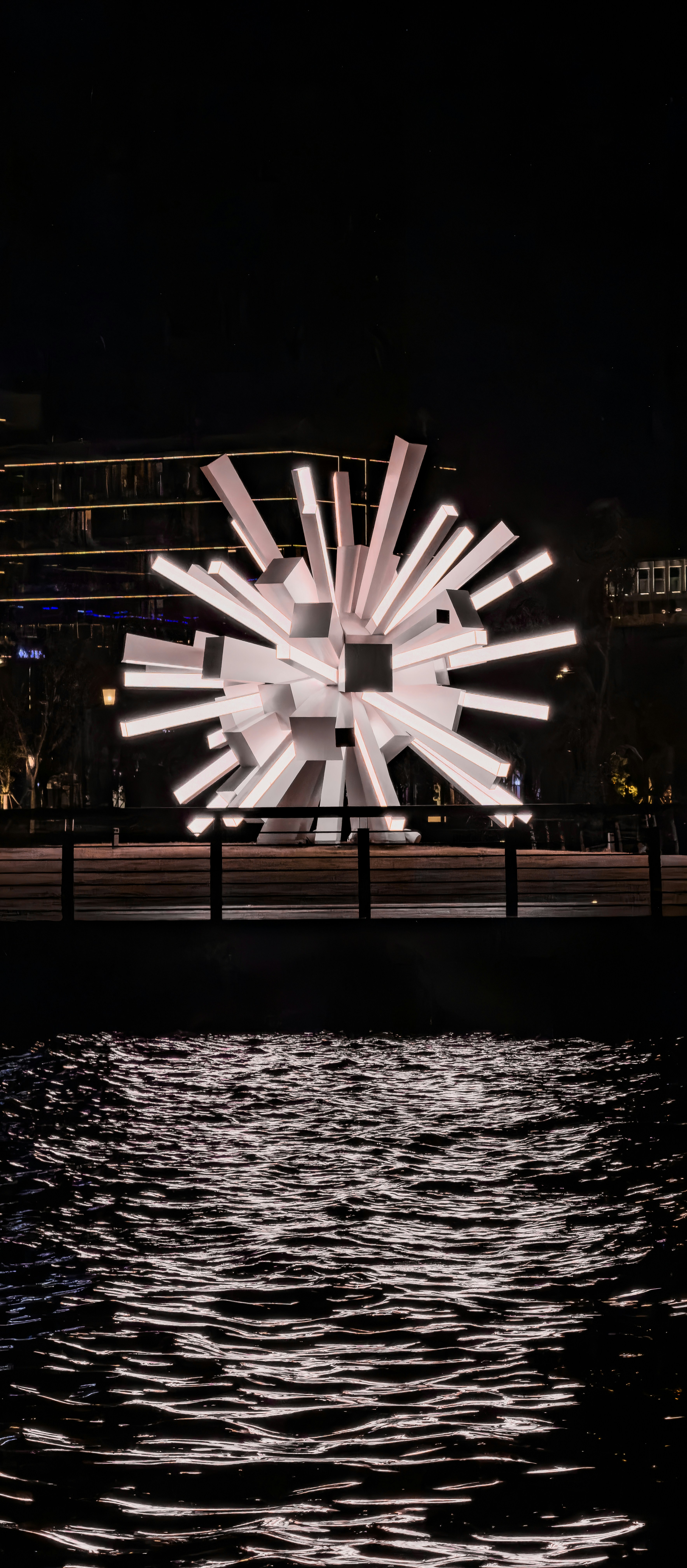 Nighttime waterfront with a glowing radial sculpture as the focal point; calm water reflects the sculpture’s light and a dark urban backdrop.