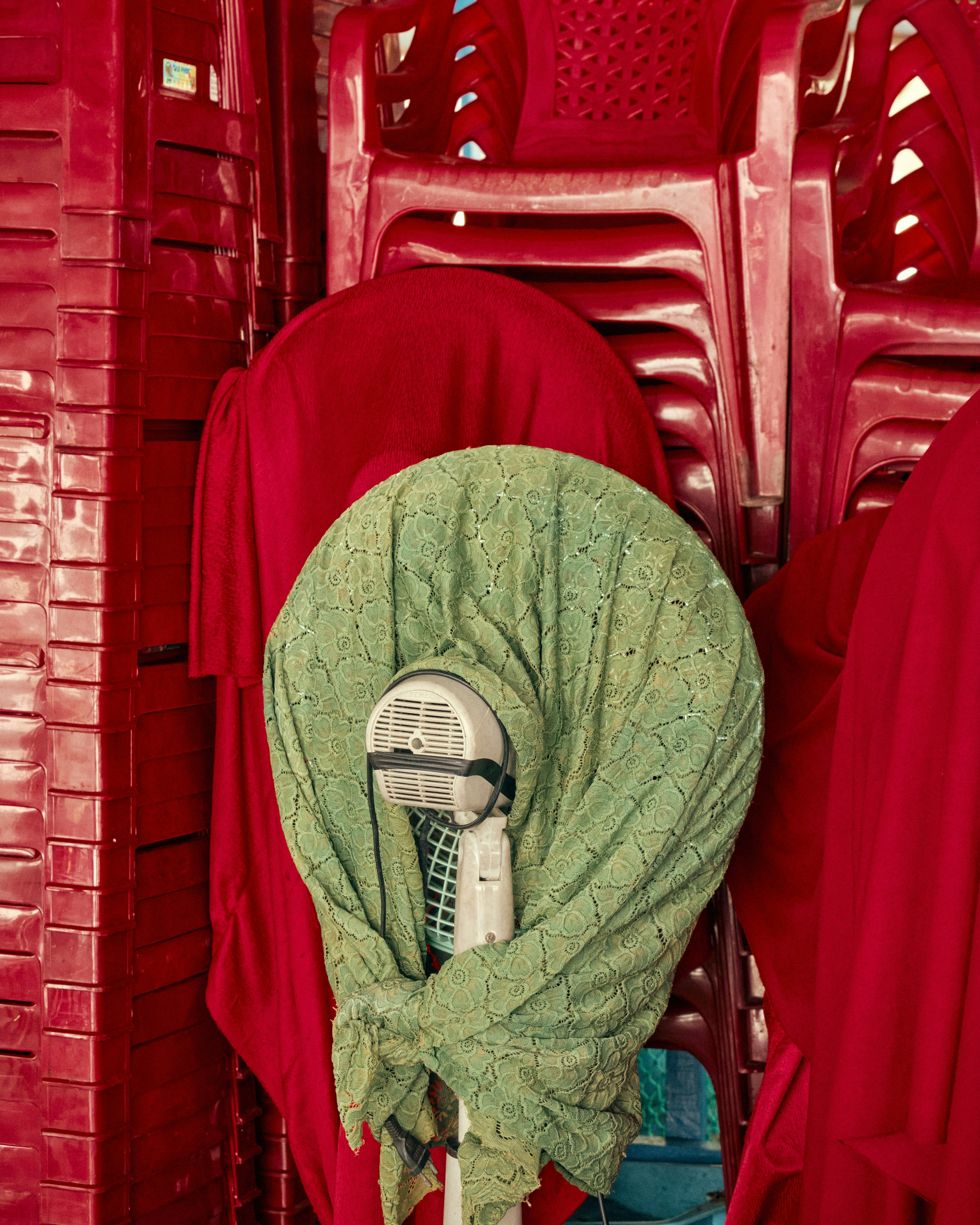 Green quilted hood drapes over a white handheld fan, set against a backdrop of stacked red plastic chairs and a red curtain.