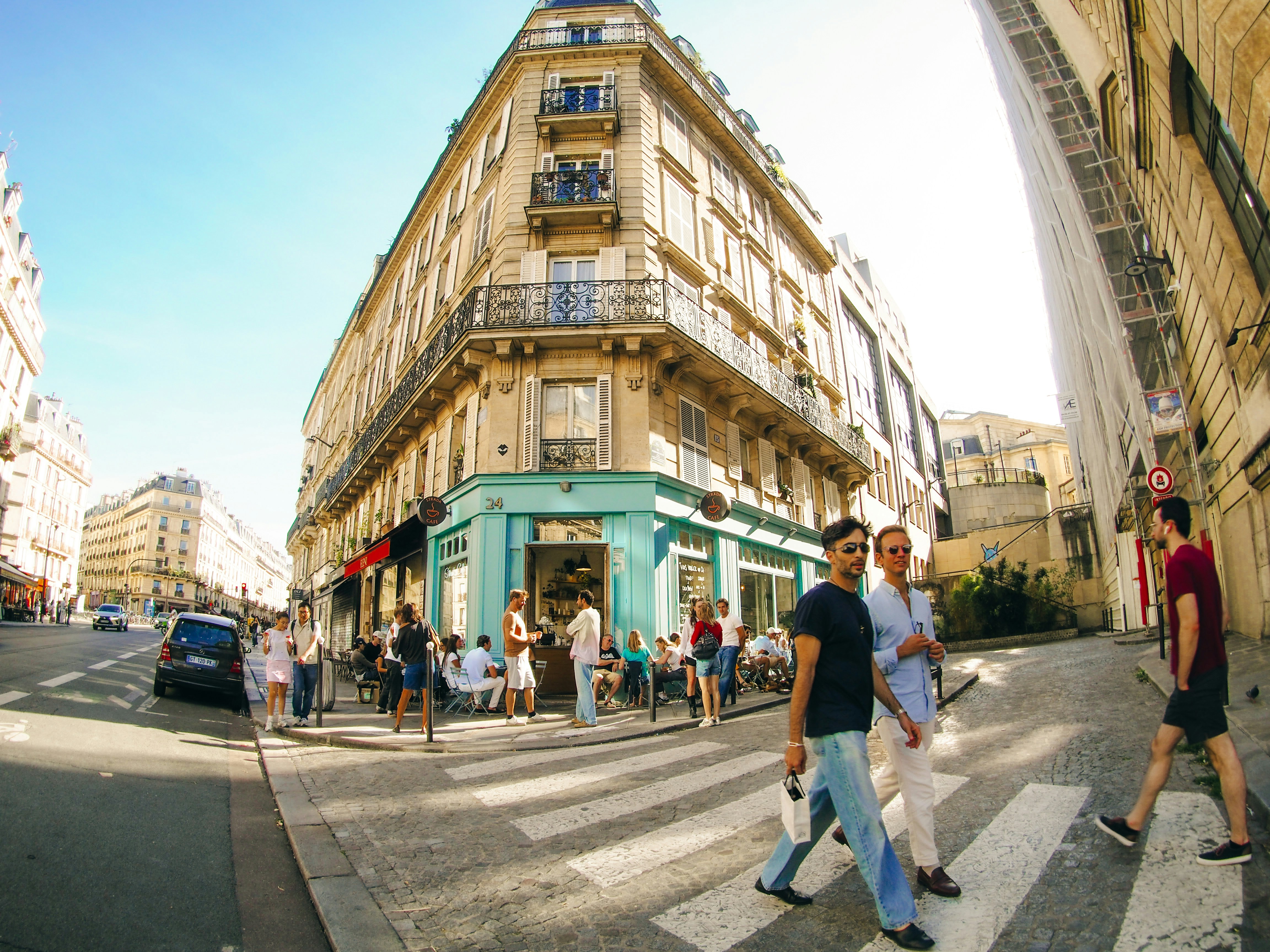 Two men walk casually past a charming Parisian café, captured with an ultra-wide 9mm lens that opens the scene into a dynamic piece of urban street photography. The wide perspective emphasizes the architecture, sidewalk, café atmosphere, pedestrians, and surrounding Parisian streets while keeping the two figures naturally integrated into the city. Warm daylight creates beautiful contrast across the historic facade, pavement, windows, and café exterior. The photograph captures authentic everyday life in Paris, combining travel, architecture, lifestyle, fashion, friendship, movement, and spontan