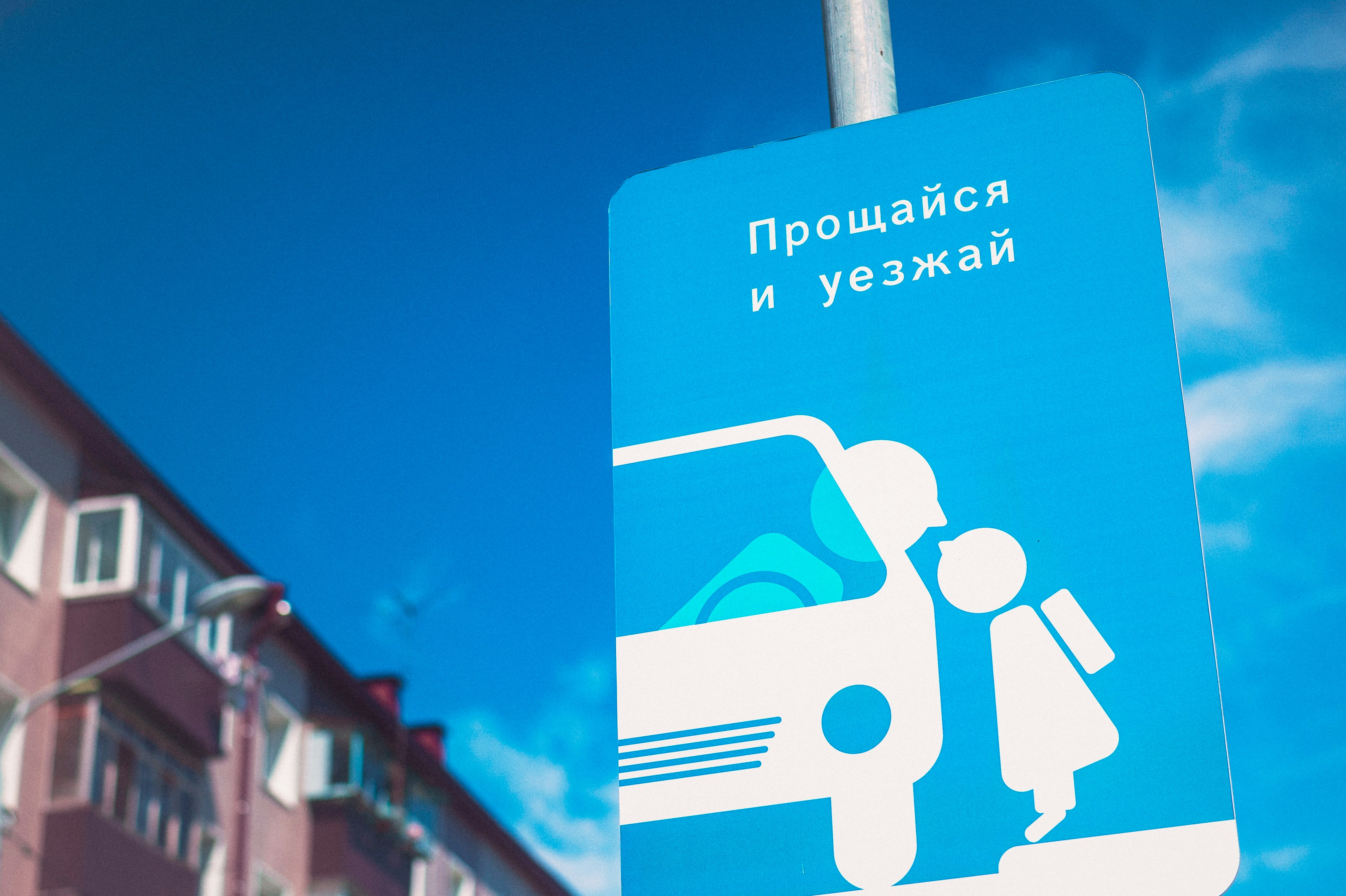 Blue pedestrian sign with a stylized figure near a building; clear sky forms backdrop, indicating a crosswalk or caution area in an urban setting.