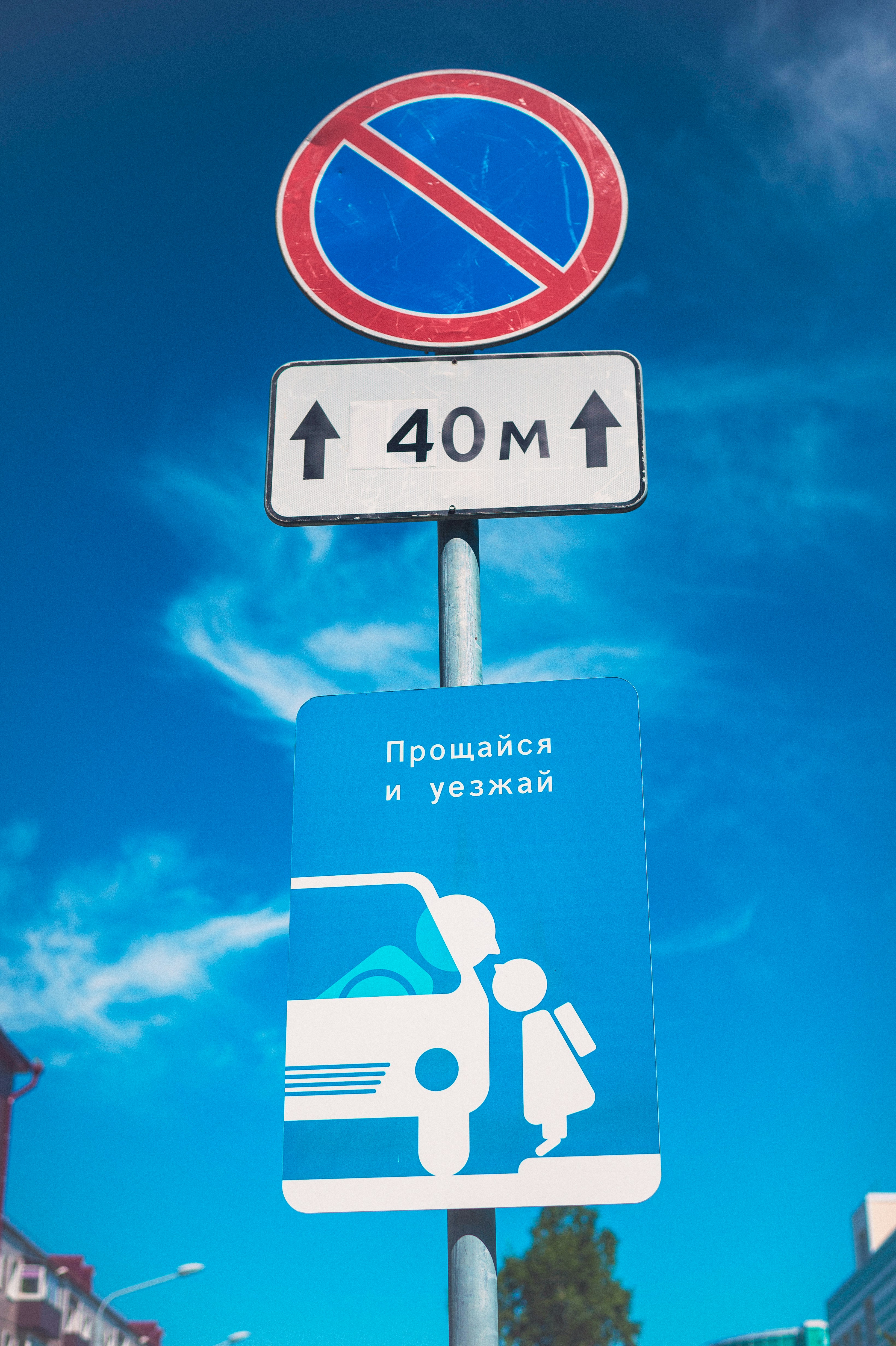 A bright, graphic street sign in Sakhalin stands out against a deep blue sky: a round no-parking symbol and a 40 m direction plate sit above a bold blue panel showing a parent saying goodbye to a child. The Russian text reads “Прощайся и уезжай” (“Say goodbye and go”), a simple, human reminder captured as clean, colorful urban signage and travel photography.