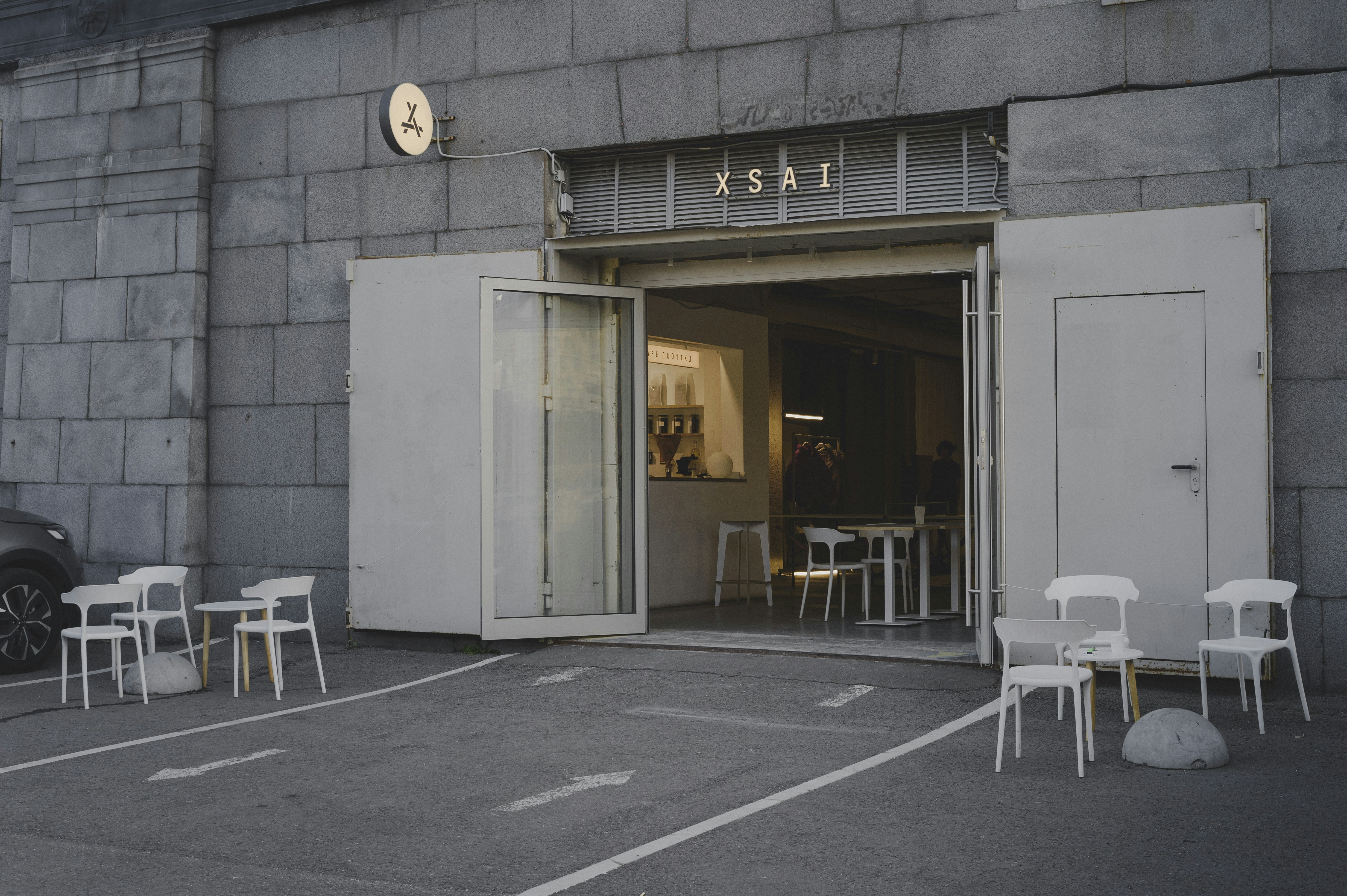 A storefront café with open doors and white chairs arranged outside, inviting a calm, unoccupied interior.