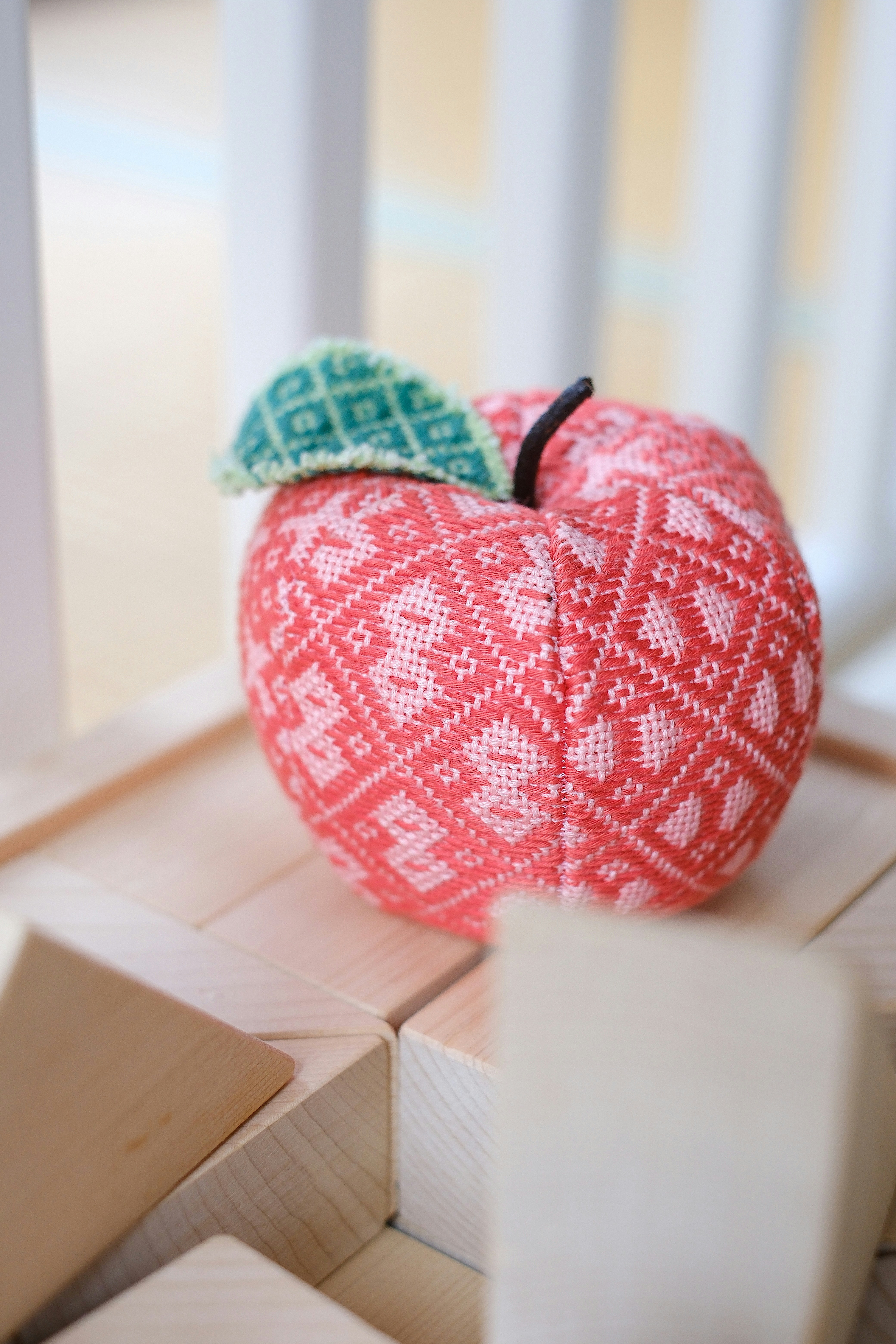 A red decorative apple with geometric white patterns sits atop light wooden blocks, with a soft-focus background and a green patterned leaf. The scene emphasizes craft textures and gentle light.