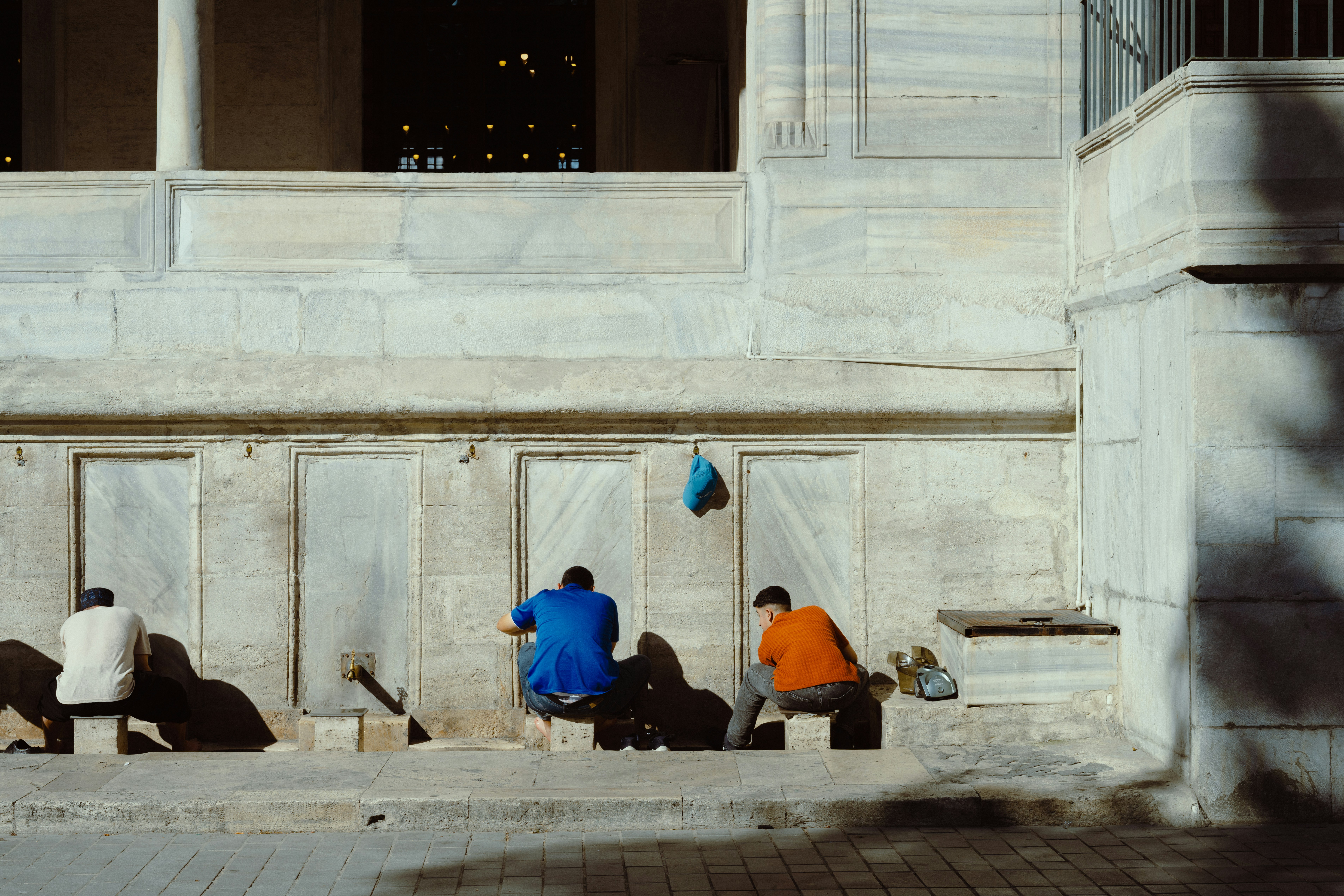 Three laborers sit against a pale stone wall with their backs to the camera; a blue bag hangs above while a bright orange shirt catches the eye, suggesting a moment of pause in a quiet architectural setting.