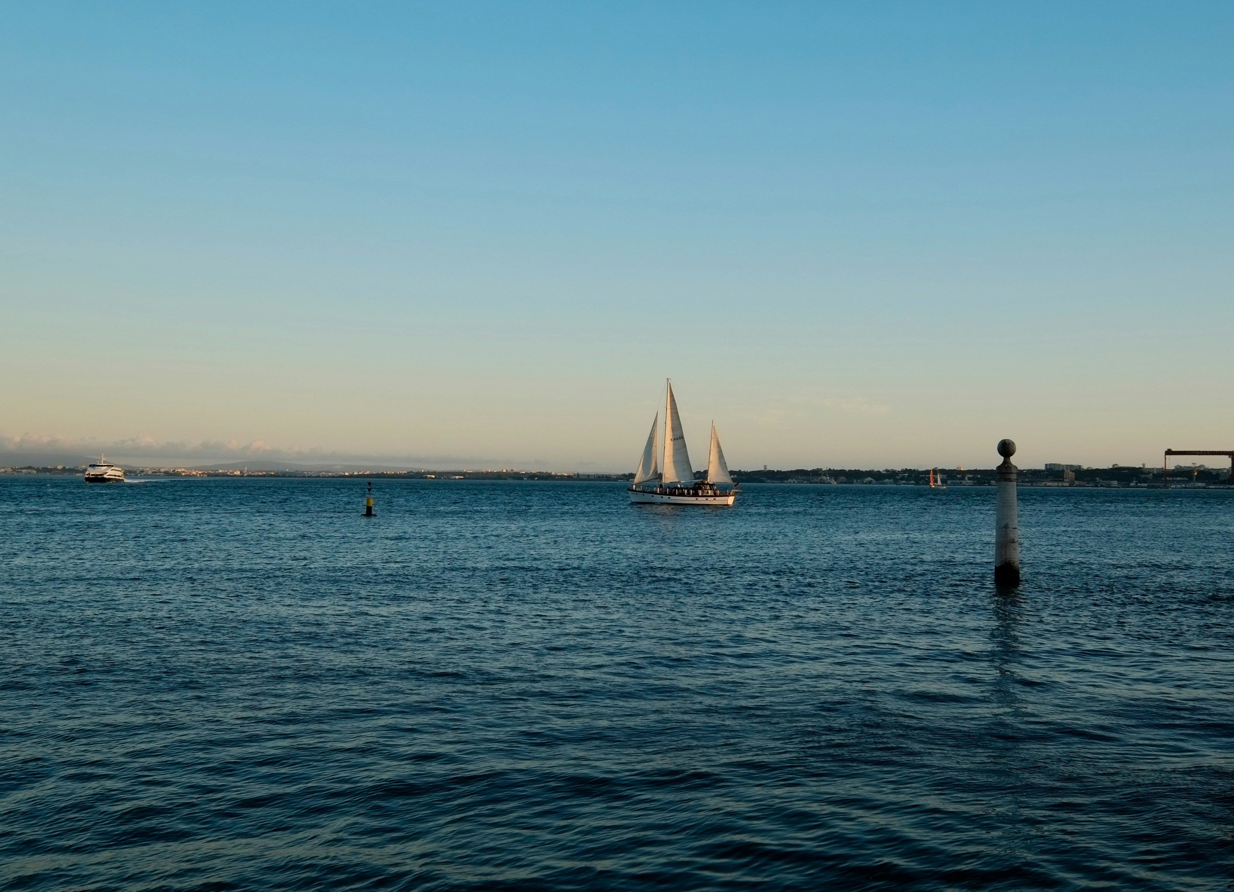 Sailboats drift across a tranquil bay with a clear blue sky and subtle sunset glow; a distant horizon anchors the scene.