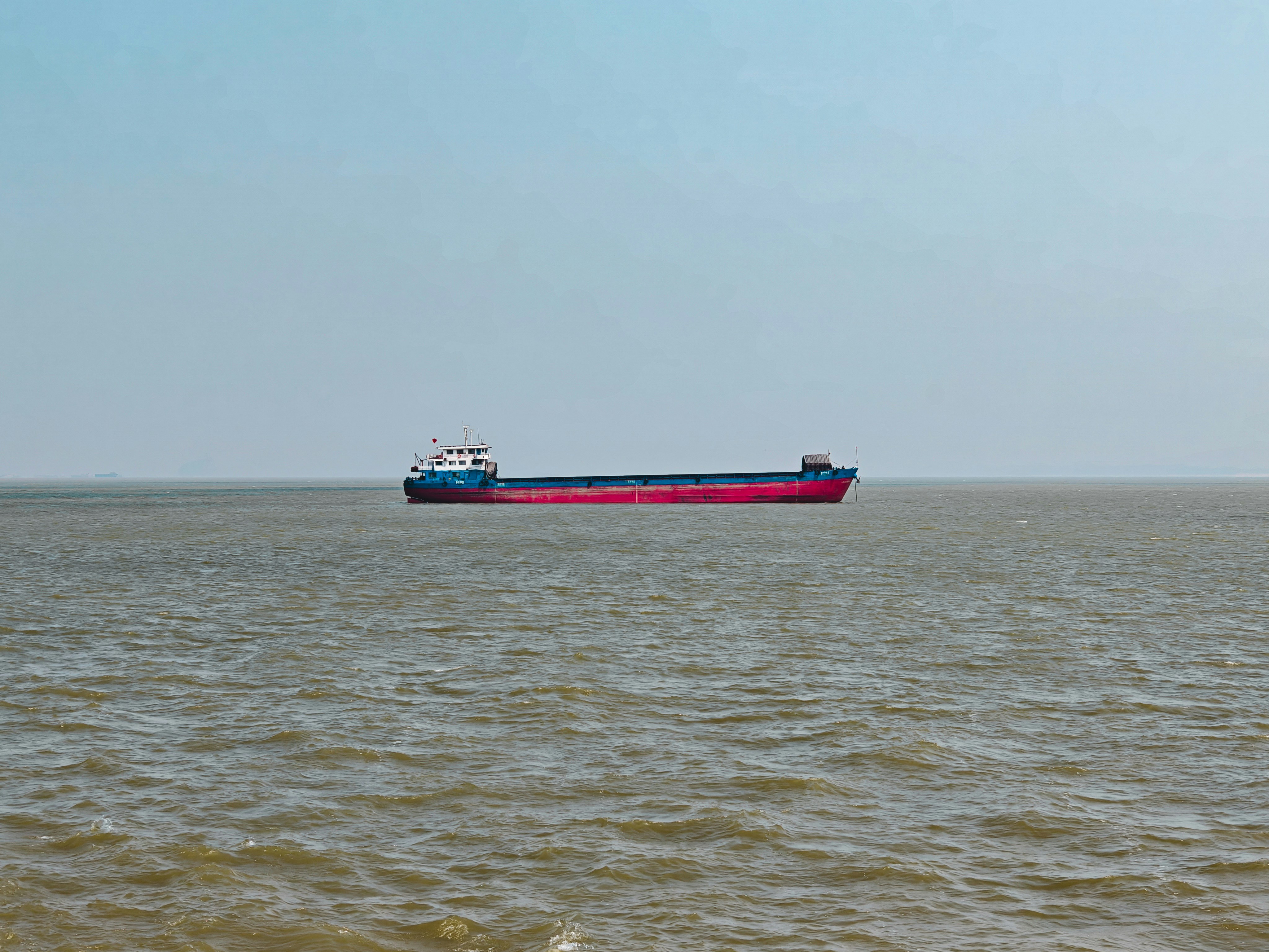 Red-tinted freighter slicing a calm sea toward a pale horizon