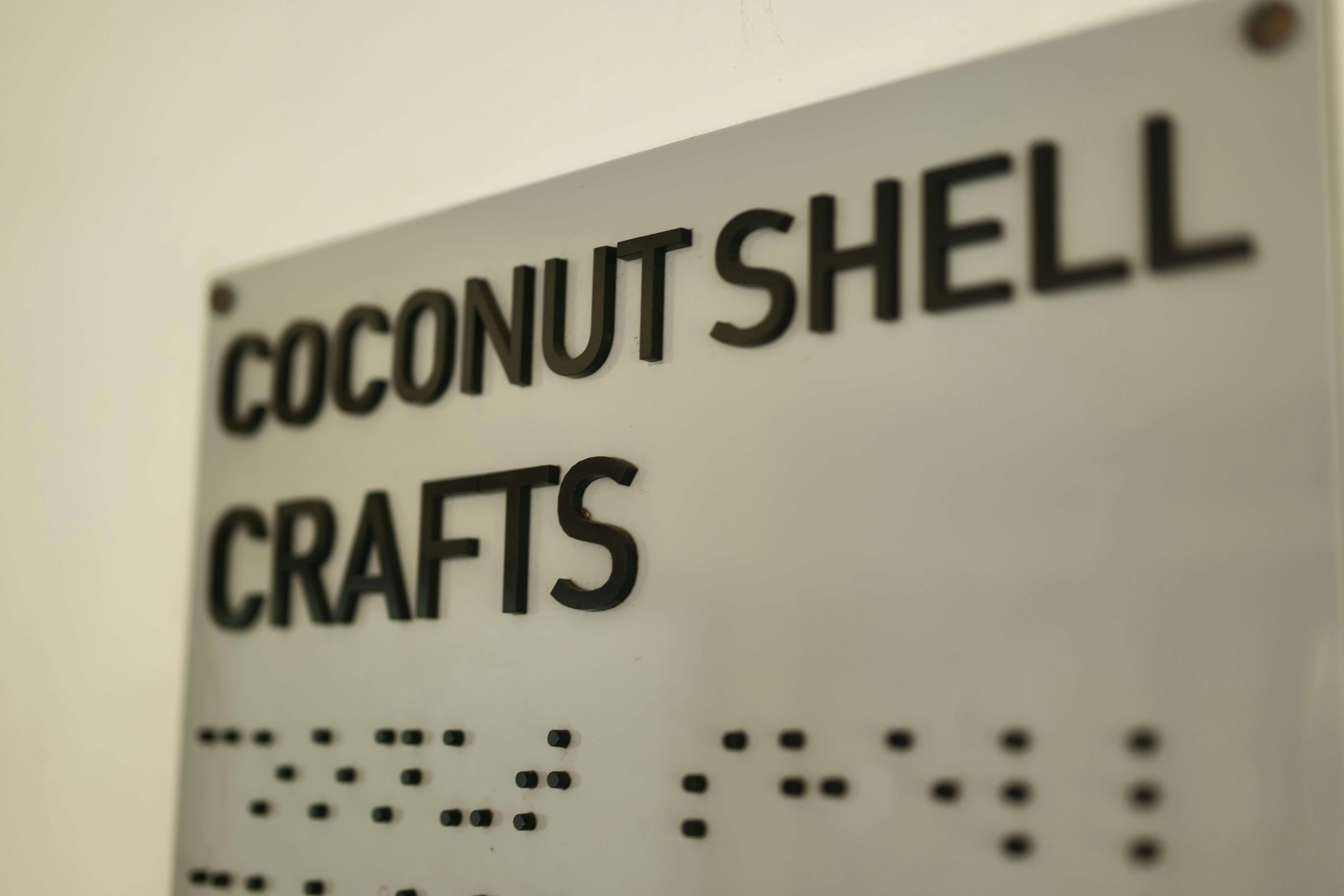 A sign reading "COCONUT SHELL CRAFTS" with black raised lettering and braille below
