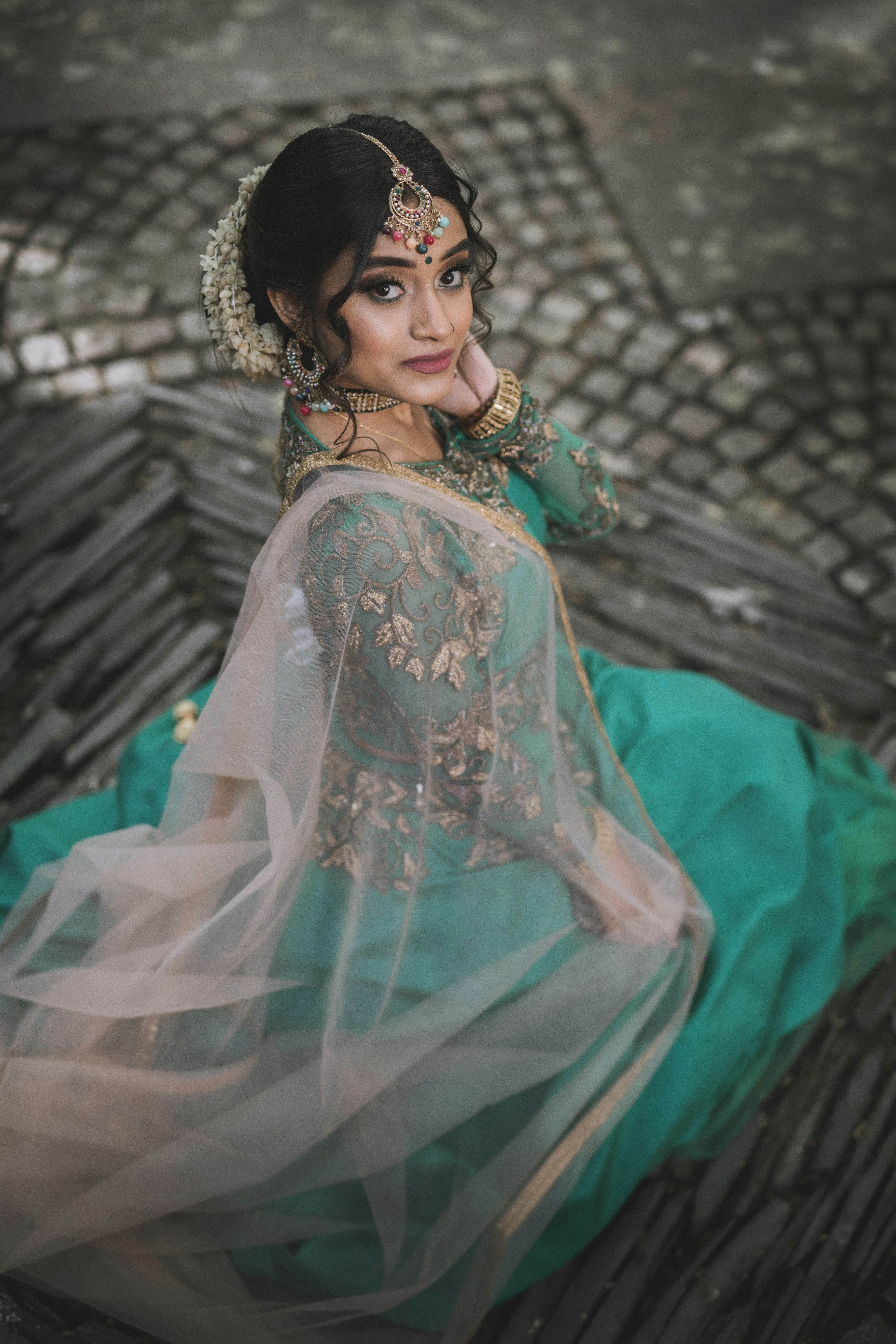 Vibrant traditional attire and jewelry adorned on a poised woman, seated on a textured stone backdrop. The portrait highlights intricate embroidery and translucent veil, emphasizing elegance and cultural detail.