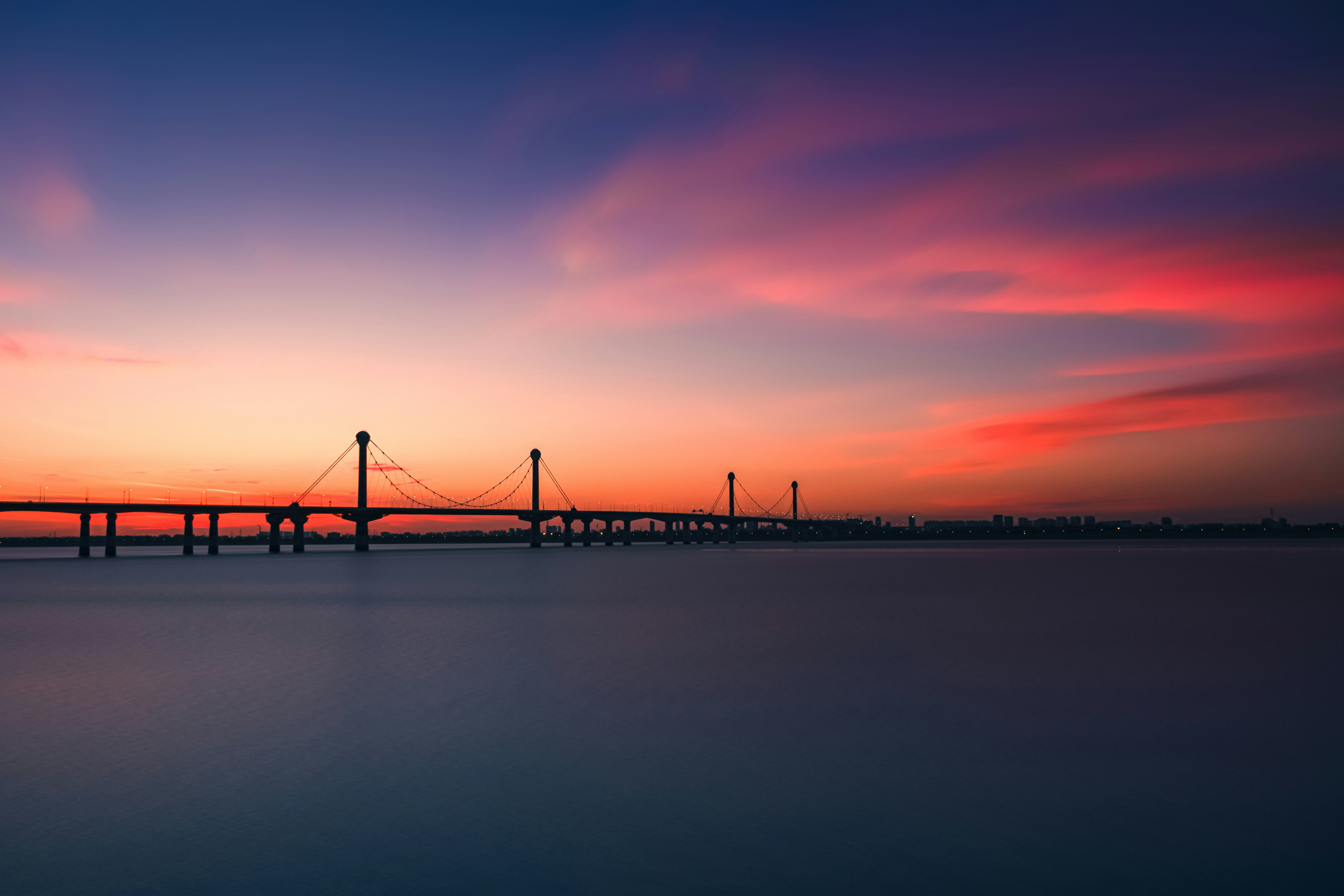 Evening sky over a calm body of water with a distant cable-stayed bridge silhouetted along the horizon, bathed in warm oranges and pinks.