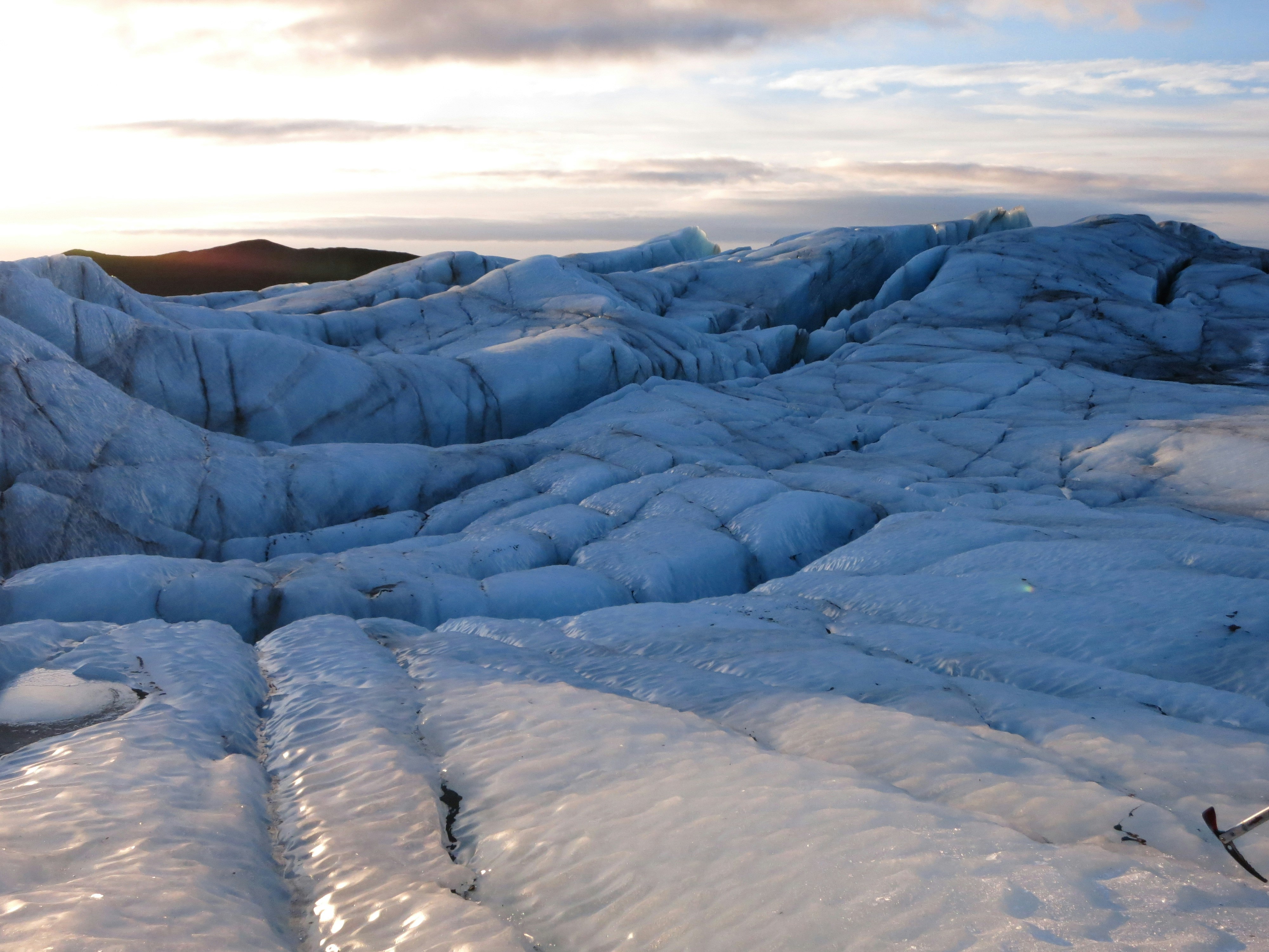 Glacial terrain with blue ice ridges stretching toward a warm sunset horizon; the uneven surface and crevasses emphasize texture and depth.