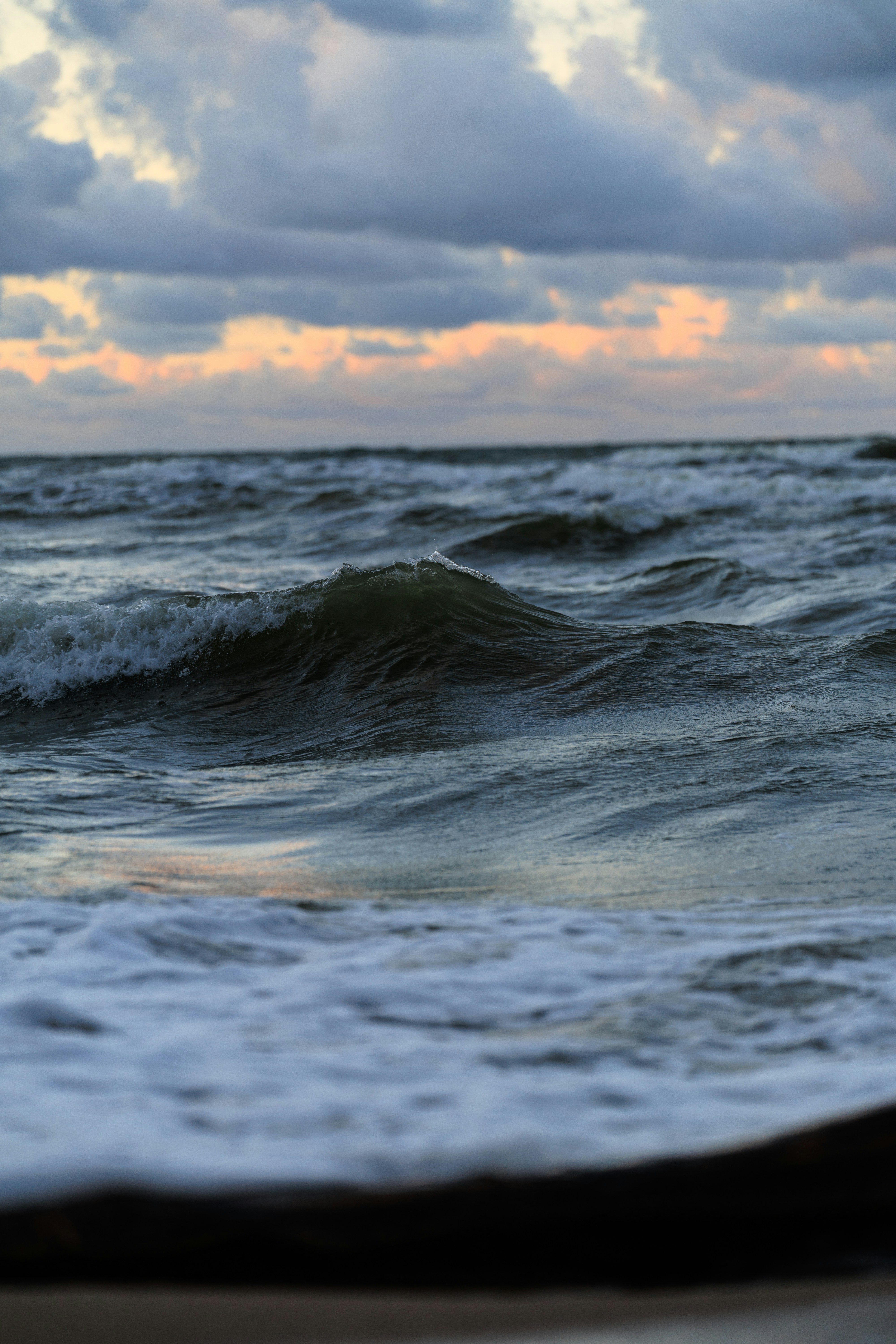 Low-angle view of rolling ocean waves approaching shore at dusk, with soft orange-hued clouds on the horizon; the scene captures the motion and texture of the sea. (Note: no logo or illustration present.)