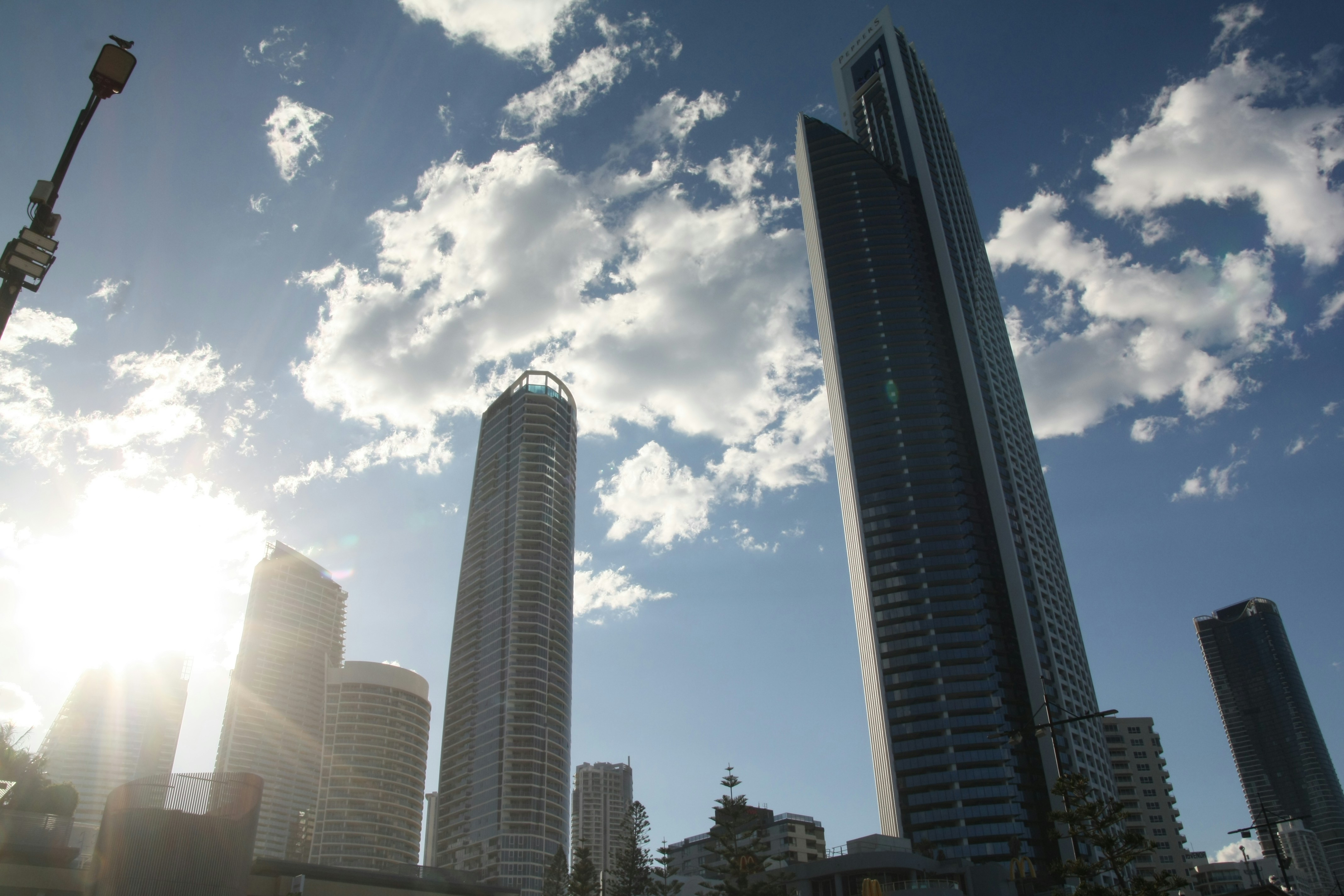 Skyscrapers rise against a bright sky with the sun flare at left; tall glass towers dominate a modern urban skyline, capturing a wide cityscape scene.